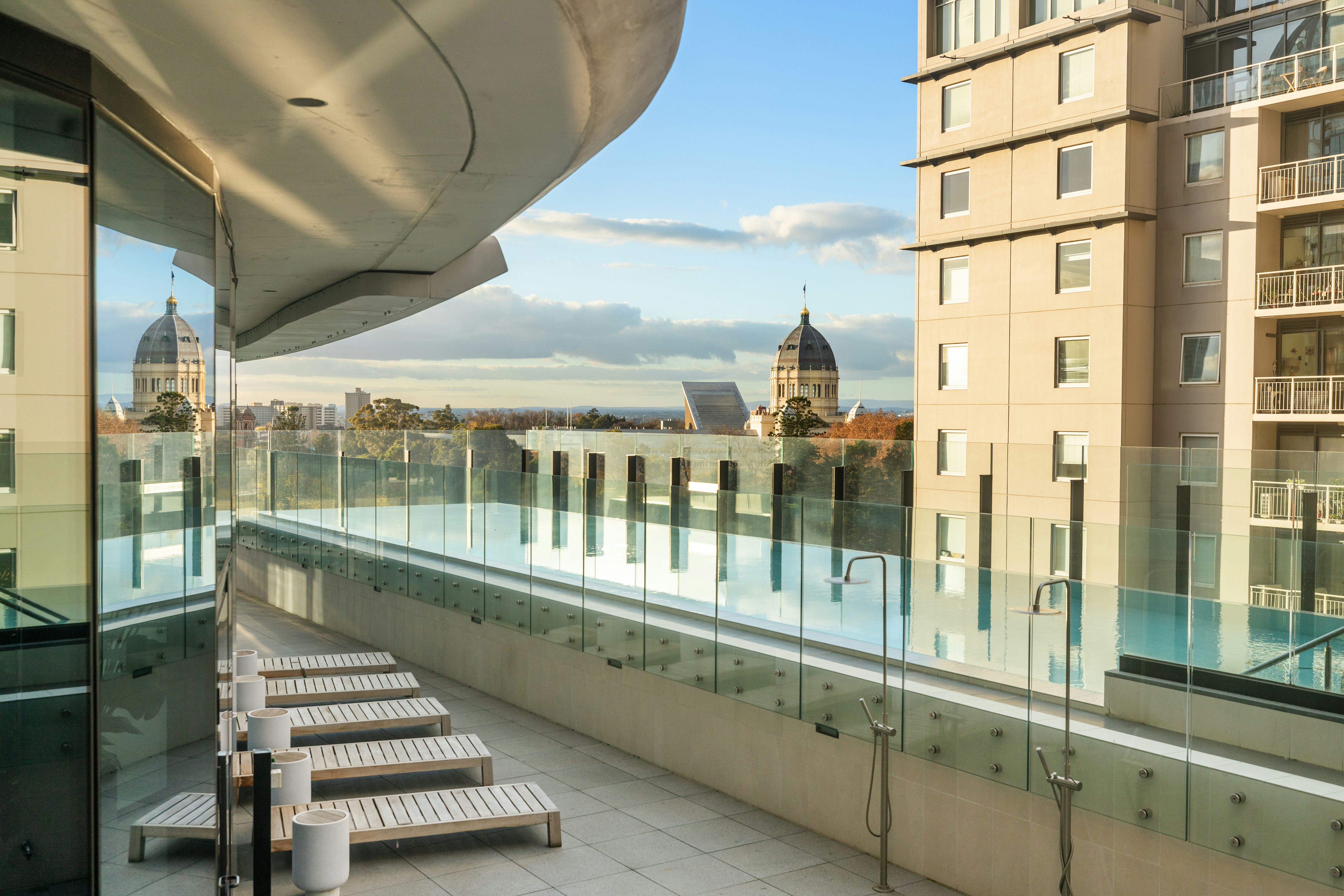 Bright terrace with glass railings overlooking city towers; lounge chairs line the left, a high-rise building on the right, and a distant skyline under a blue sky.