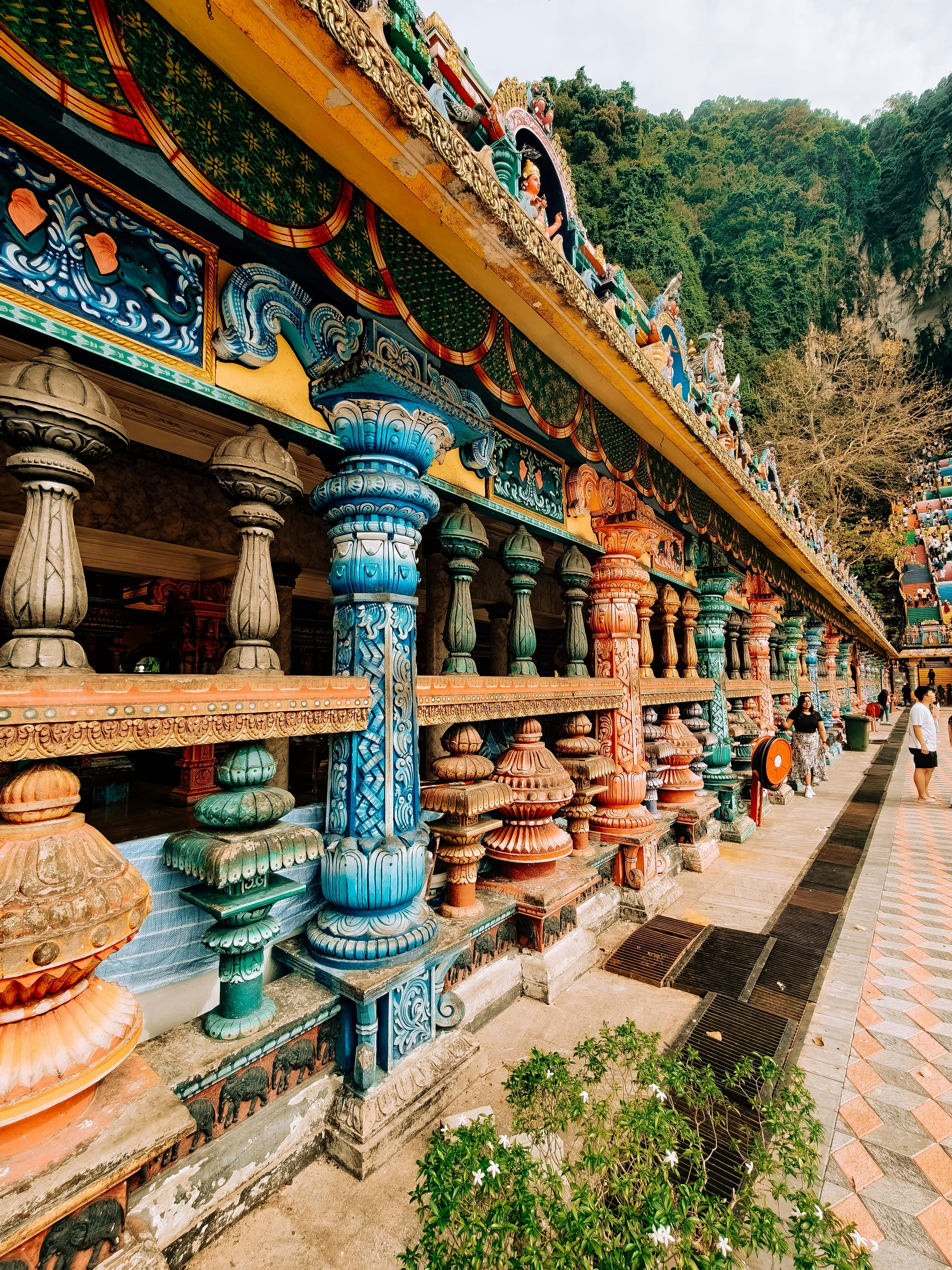 Vibrant ornately carved columns along a temple walkway, with decorative beams and a distant figure adding scale and movement.