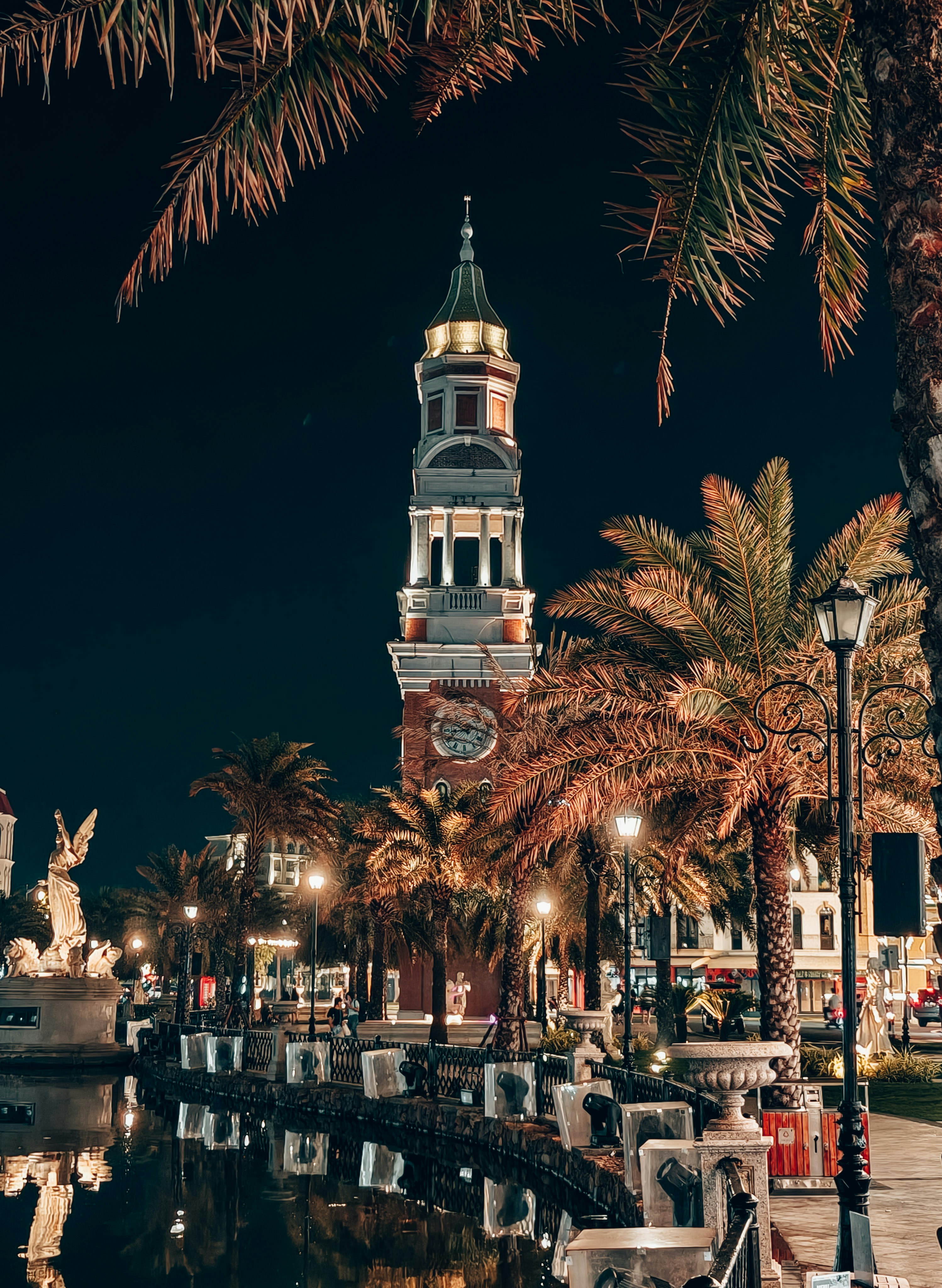 Night scene of a clock tower framed by palm trees along a lit waterfront promenade; foreground includes lampposts, benches, and a reflective canal.