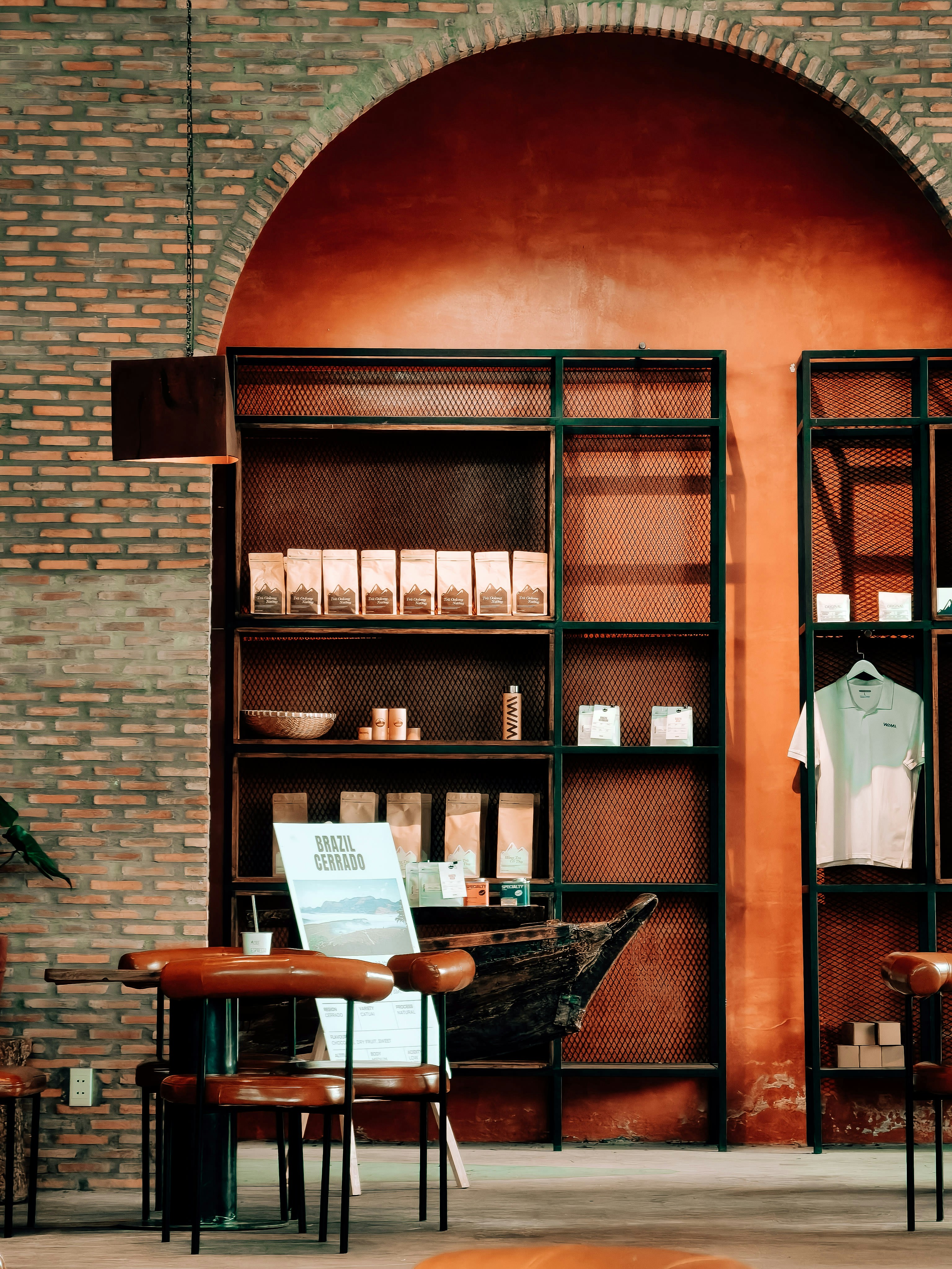 Warm-toned interior with a brick arch, metal shelving filled with products, and a small seating area; a retail display scene with clothing on the right and a central display board. Brand items and shelves suggest a cafe-like shop interior.