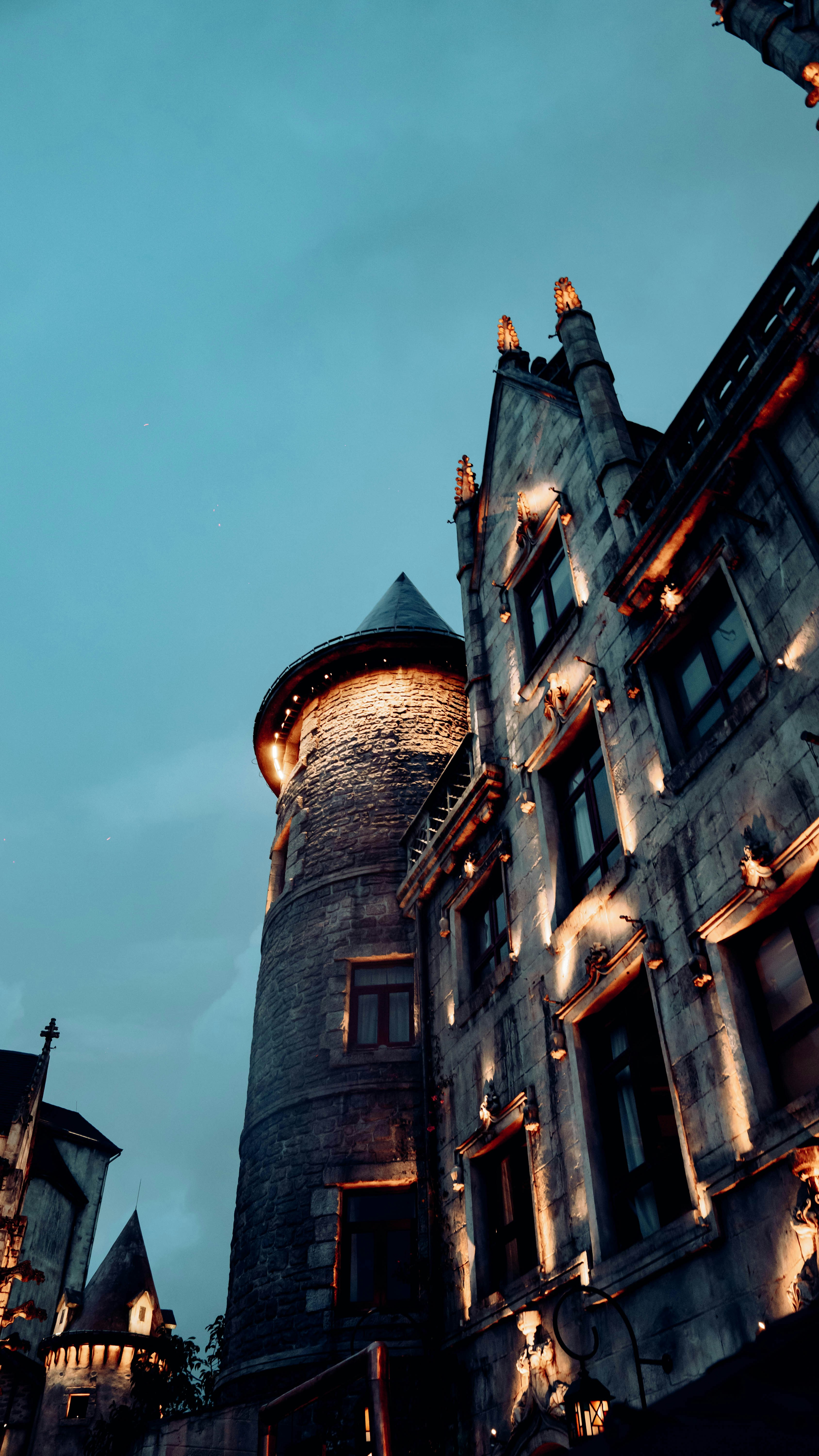 Majestic stone castle tower and illuminated facade rise against a dusky blue sky, with warm torches creating a dramatic contrast. Architectural details and texture are emphasized in this urban fantasy scene.