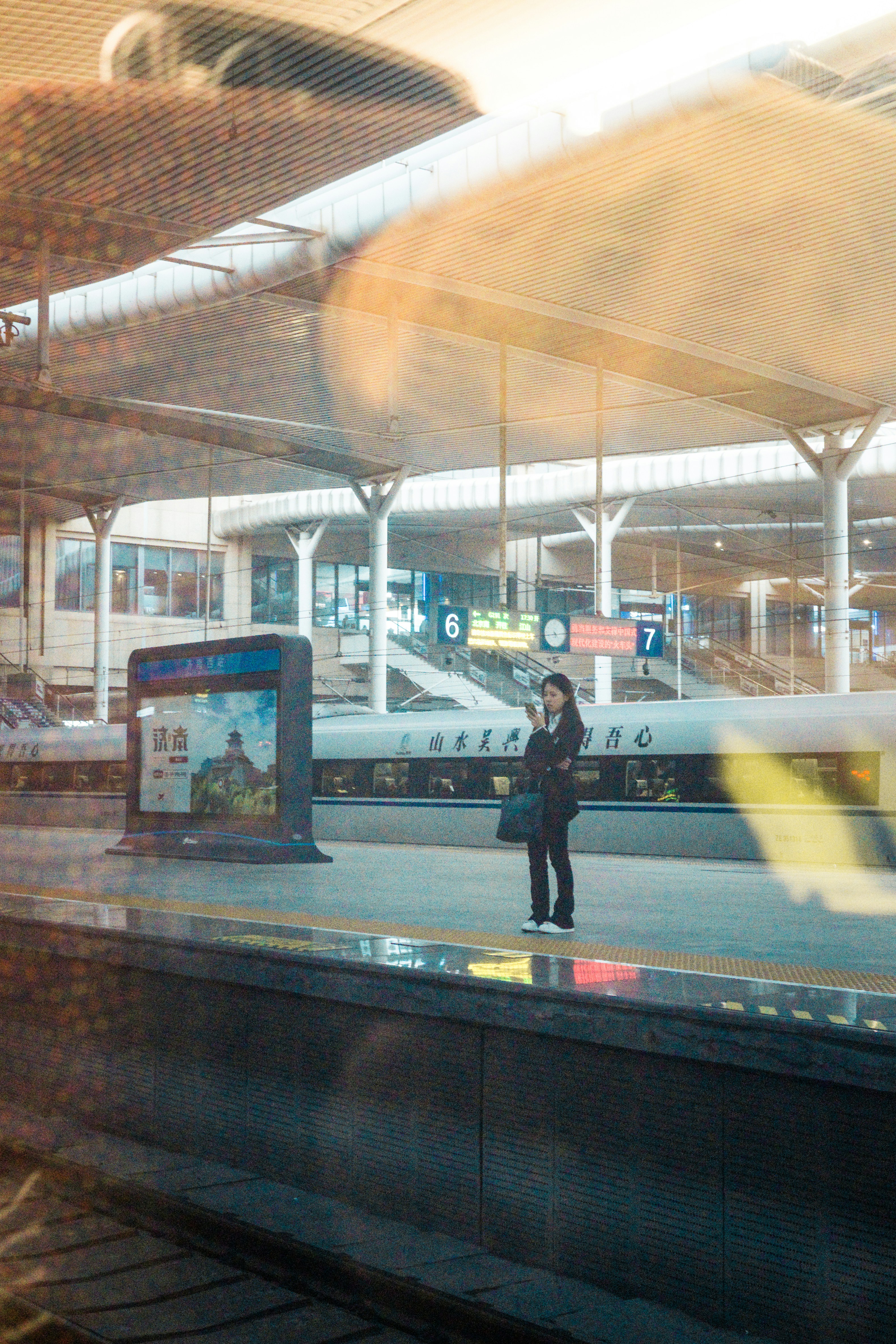 A solitary figure stands on a train platform bathed in warm light, with modern station architecture and a blurred interior reflection suggesting a window shot.