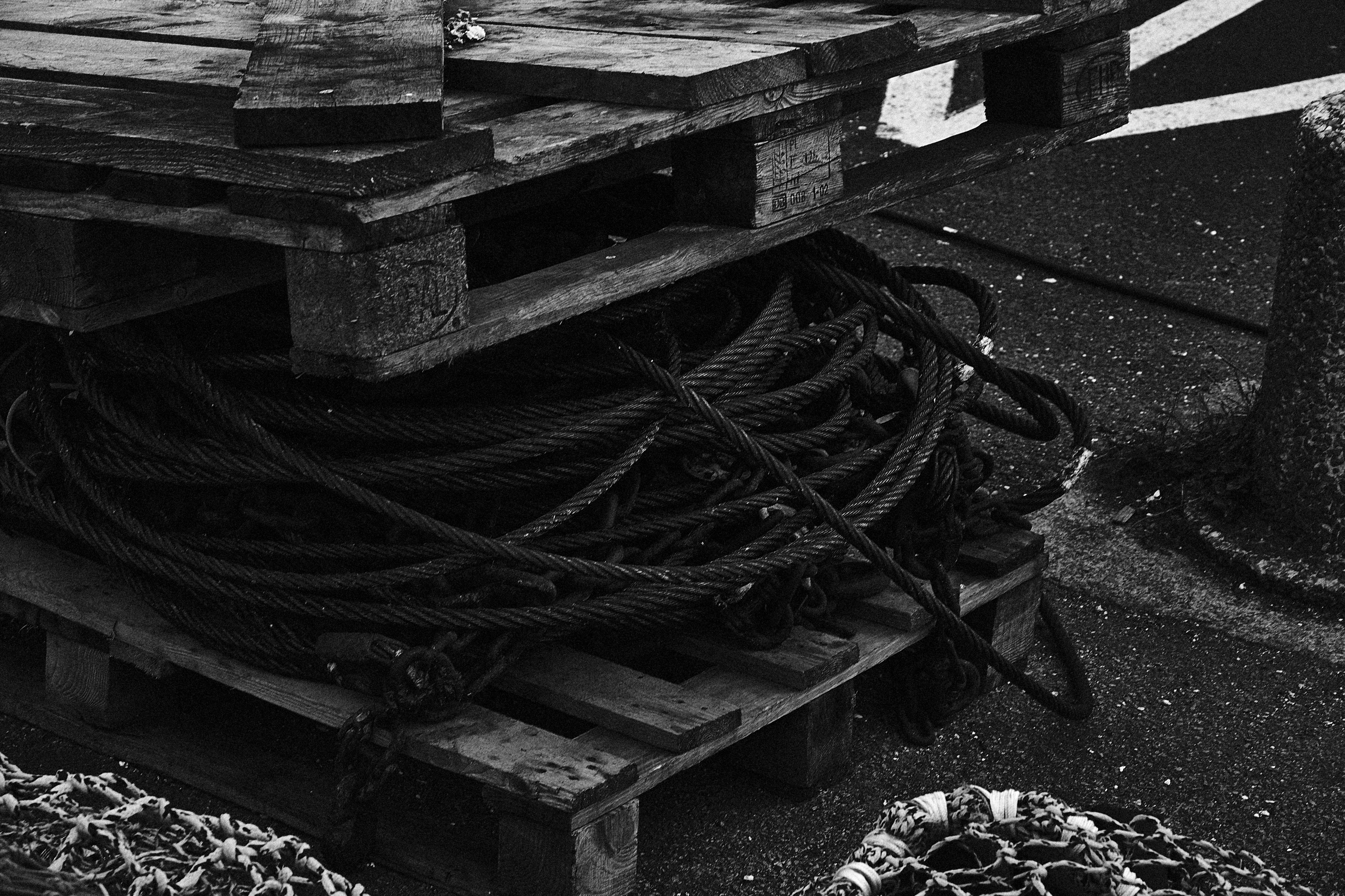 Coiled ropes and stacked wooden pallets sit on a dockside surface, with tangled lines and a gritty, industrial texture.