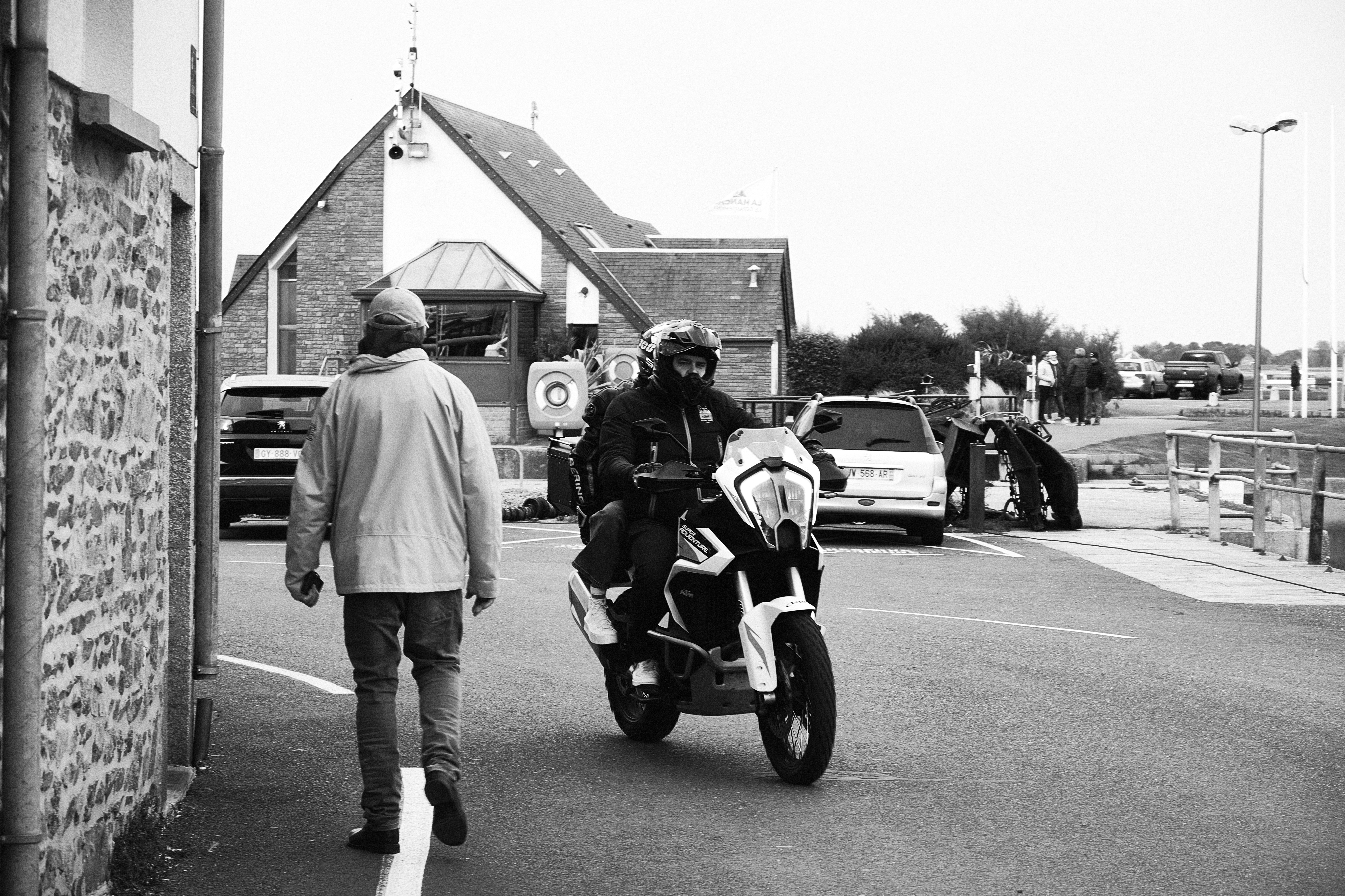 Two figures near a motorcycle in a gray-toned street scene, with a rider on a sport bike facing toward parked cars by a harbor area.