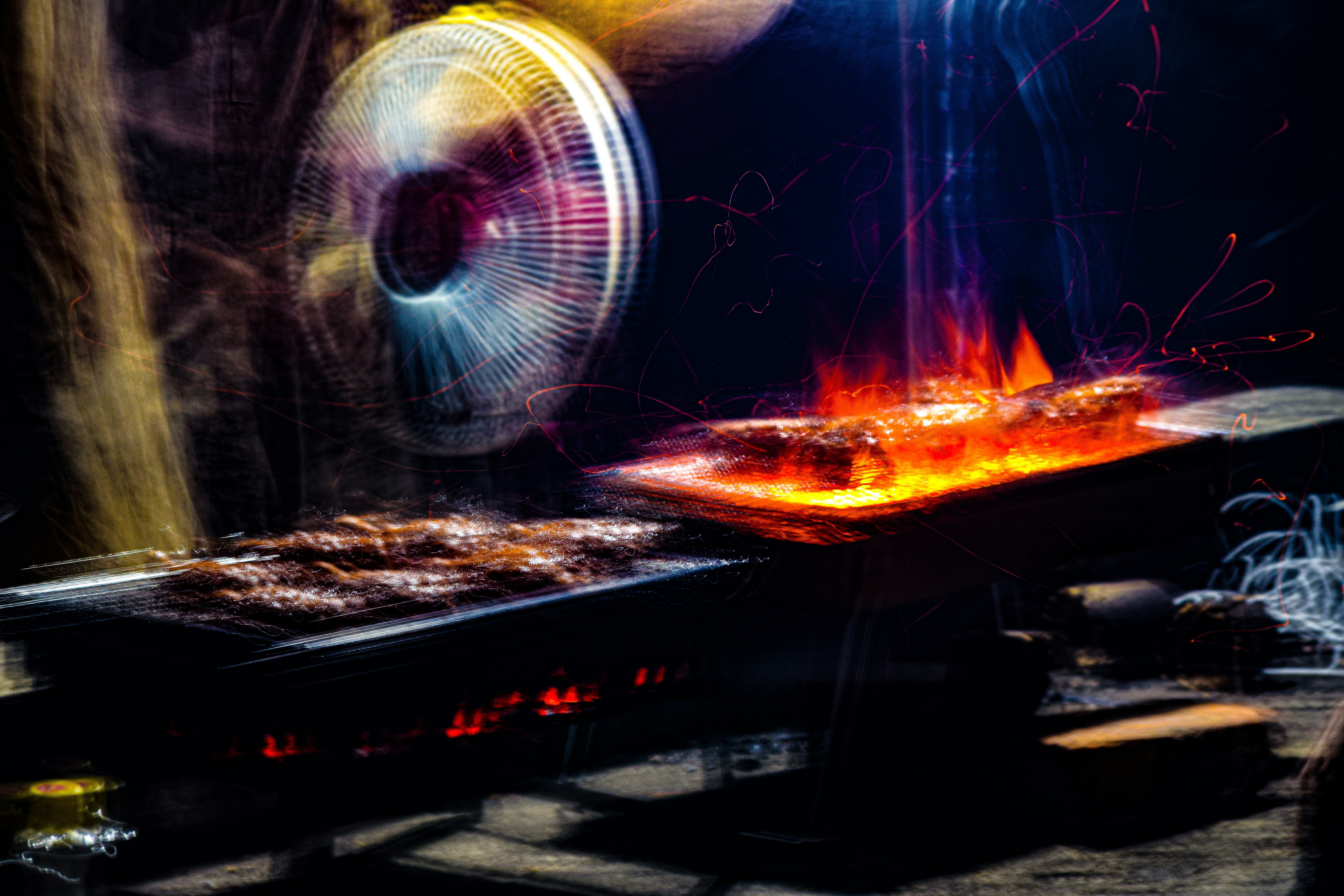 A dramatic, long-exposure snapshot capturing the raw intensity of night-time street food preparation. Fiery flames, glowing embers, and flying sparks dance across sizzling grates, while a swirling electric fan adds motion and ethereal light paths to the dark atmosphere. A striking blend of heat, smoke, and chaotic urban energy.