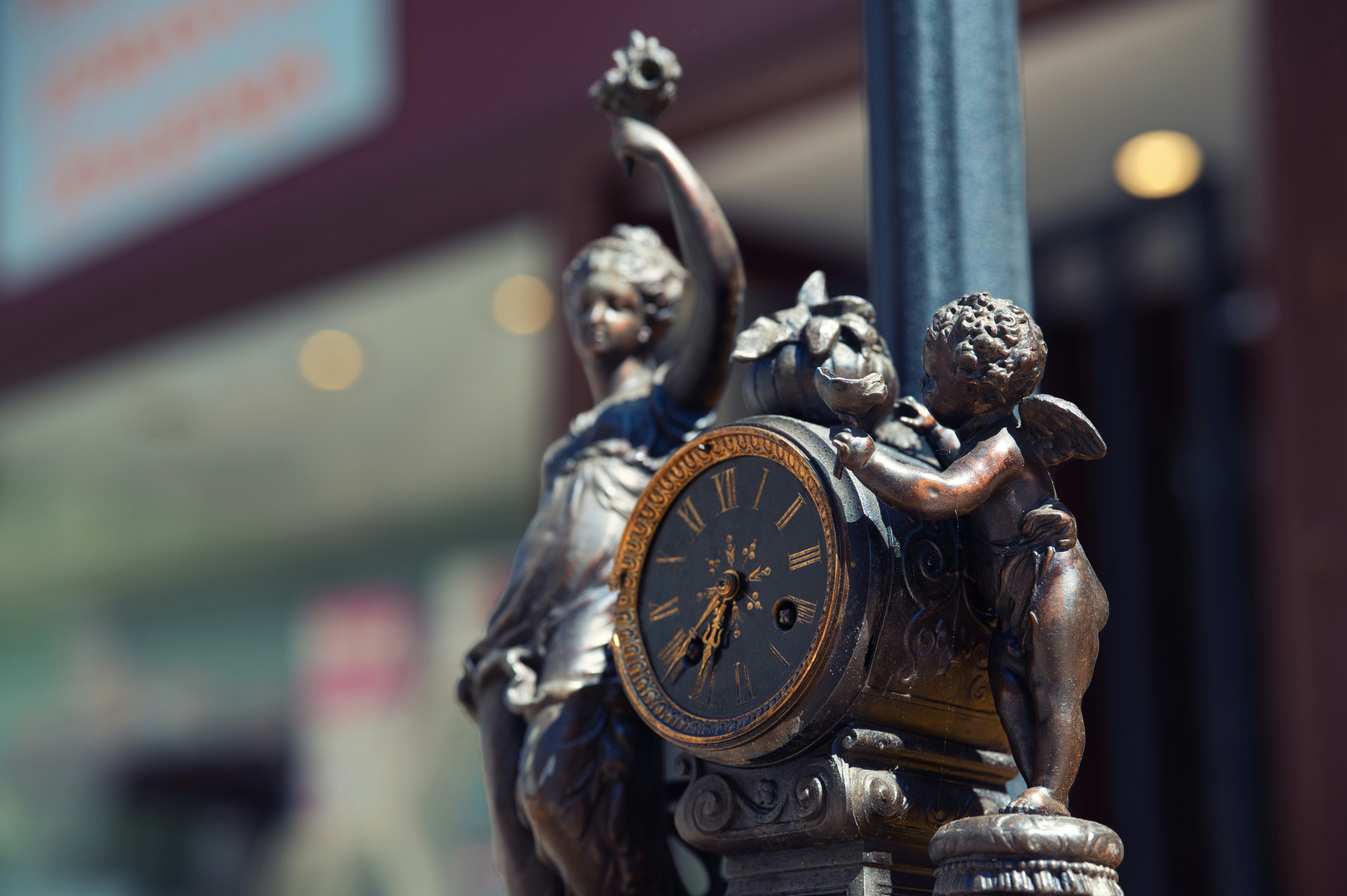Bronze clock sculpture with cherubs on a blurred urban backdrop, highlighting ornate details and weathered patina.