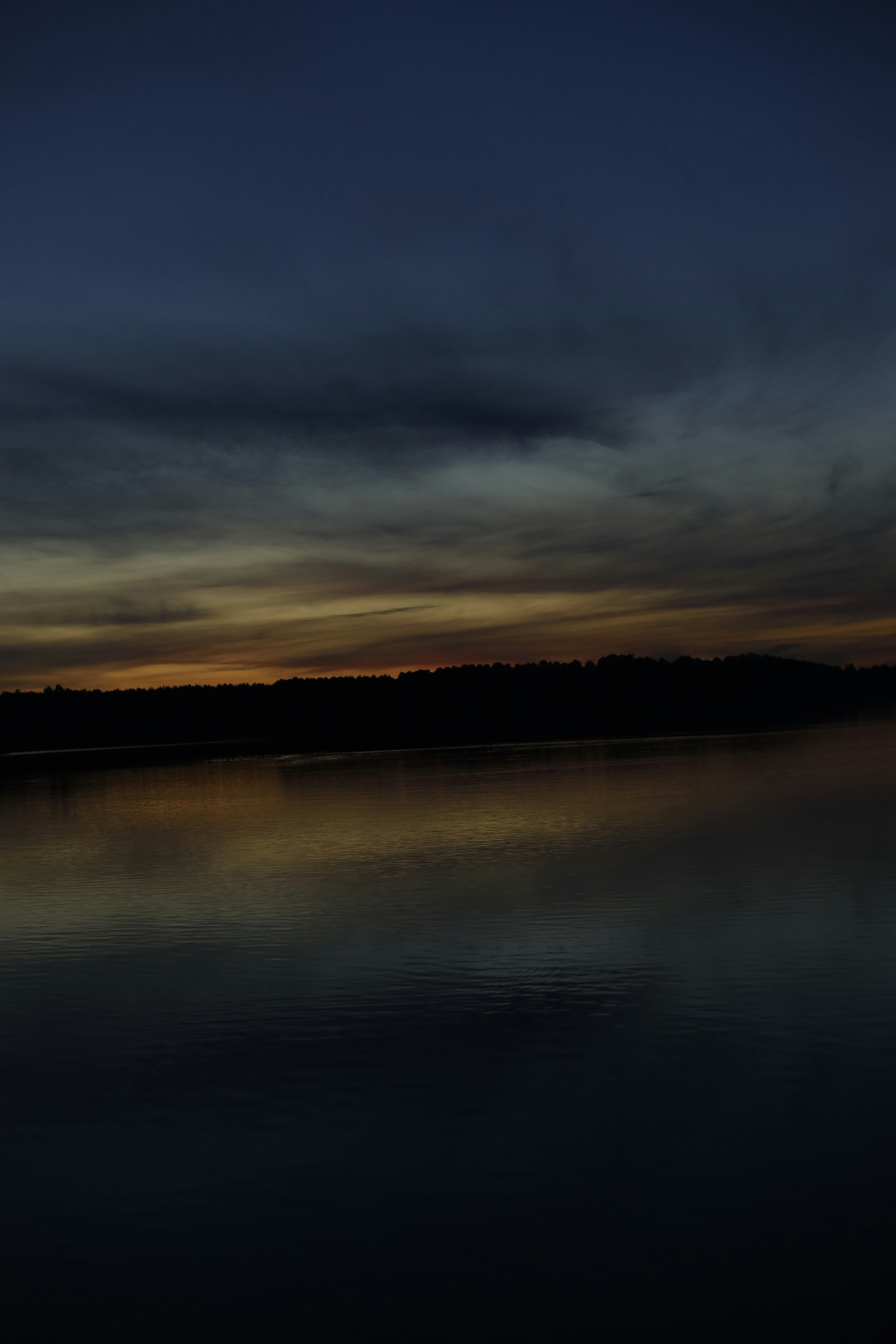 Calm lake reflects a gradient sunset with a dark shoreline; the scene focuses on the tranquil water and low-contrast horizon. This photograph is a natural landscape capture of a lakeside at dusk.