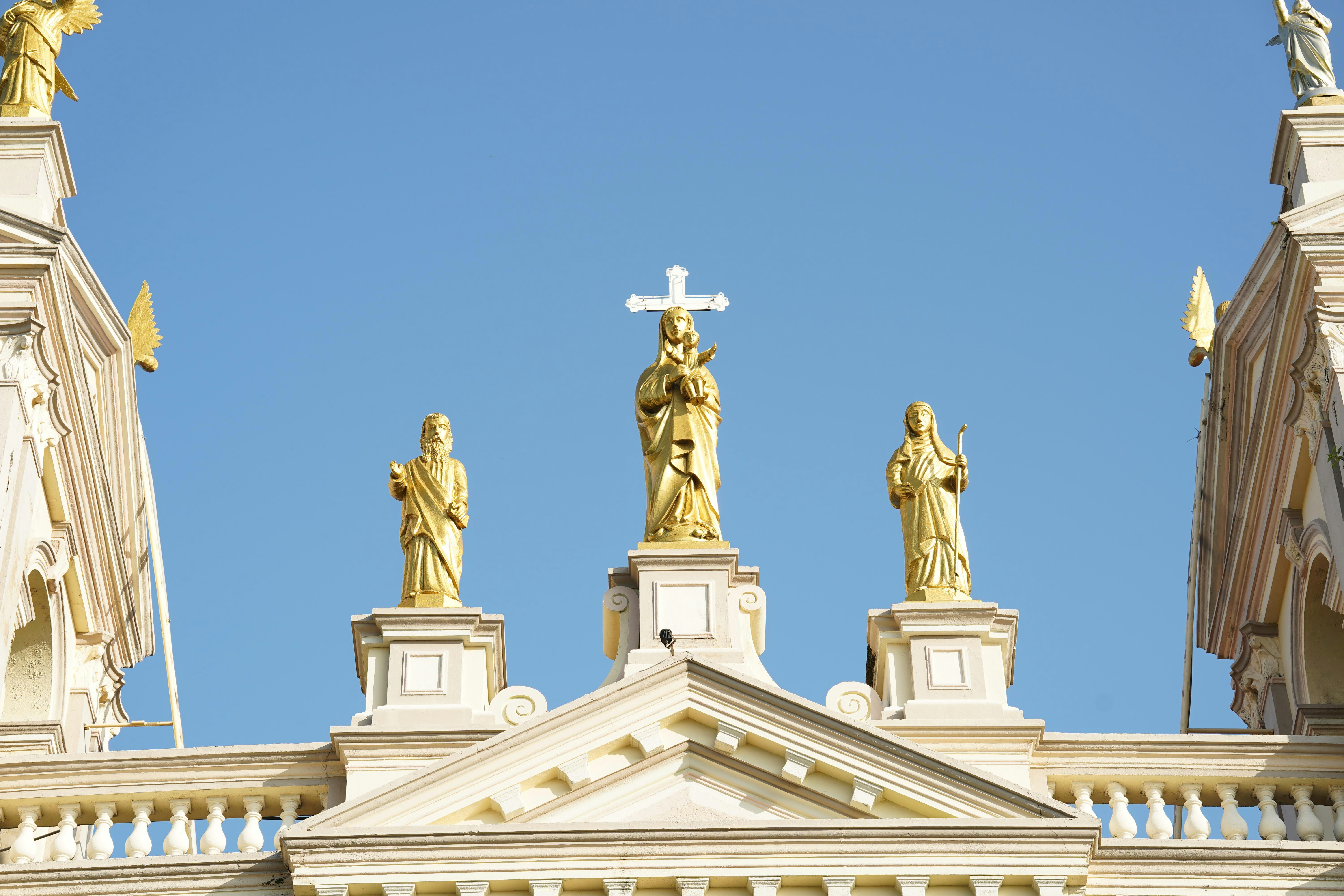 Ornate statues atop a classical pediment against a blue sky; architectural grandeur with golden figures flanking a central cross. (Logo/architecture photography).