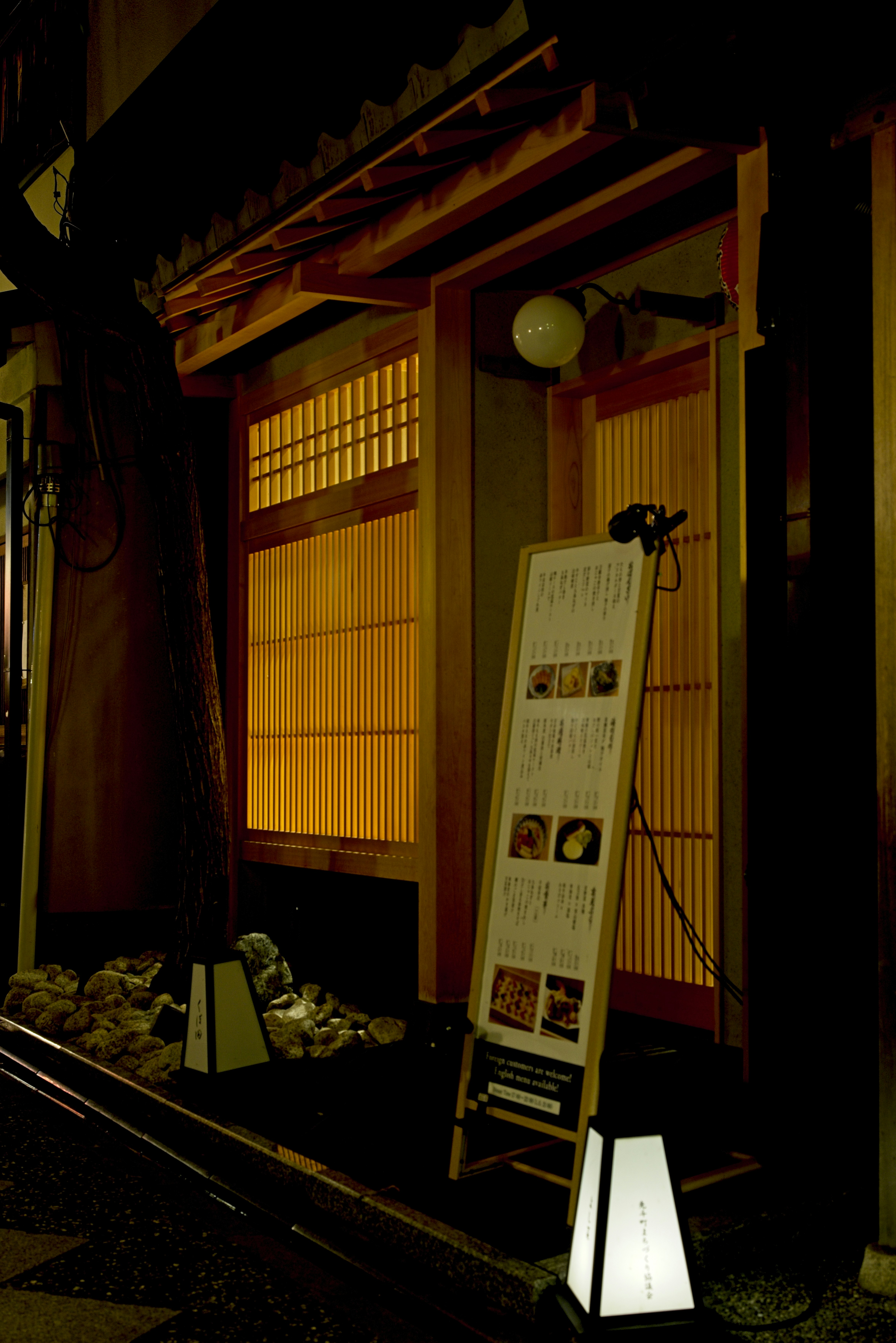 Glowing shoji screens spill warm amber light through a traditional restaurant entrance in Pontocho Alley. A tall wooden menu board displays photos of seasonal dishes beside the softly lit paper globe lamp above the doorway. Small stone triangular signs and a scattering of pale rocks line the threshold, hinting at a tucked-away eatery welcoming guests in for the evening. The humid July night settles over the narrow lane, the wood and paper textures catching the restaurant's inviting glow — a quintessential scene of old Kyoto hospitality after dark.