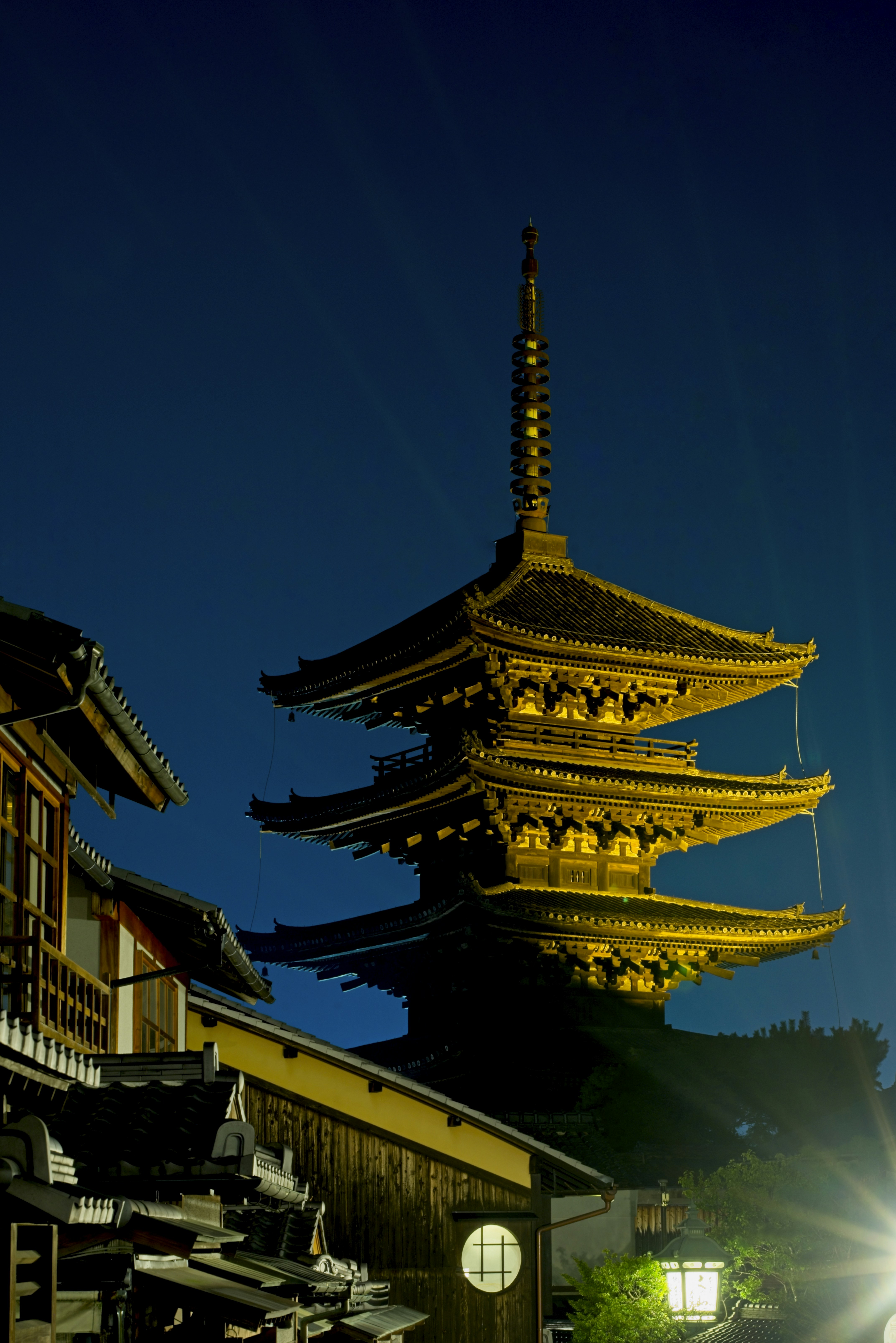 The golden-lit tiers of the Yasaka Pagoda at Hōkan-ji Temple rise dramatically above traditional wooden machiya rooftops in Kyoto's Higashiyama district on a humid July evening. Illuminated lanterns and a round latticed window glow softly at street level, framing the pagoda's intricate bracket work and towering sōrin finial against the deep blue night sky. This striking composition captures the enduring architectural elegance of one of Kyoto's most photographed landmarks.