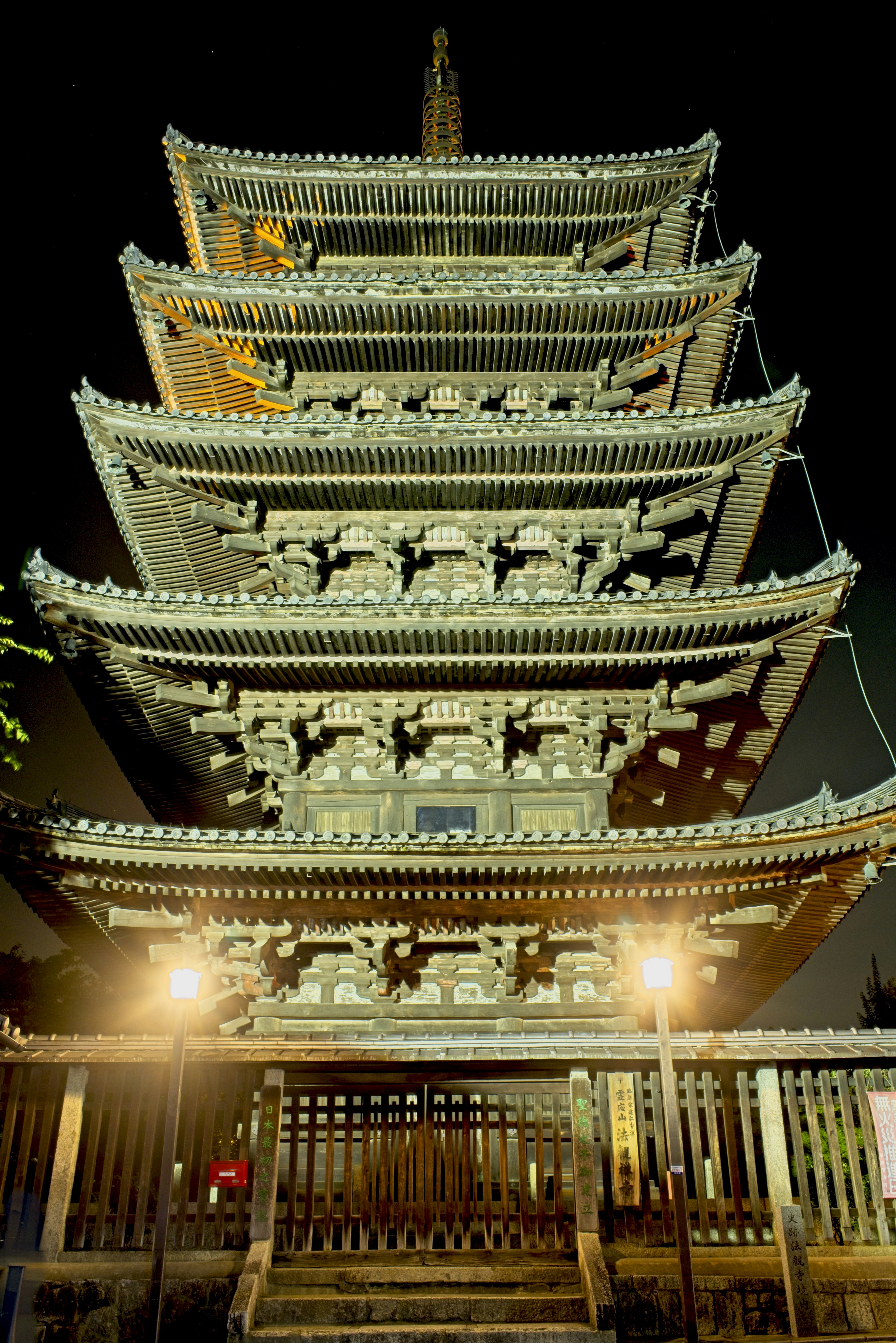 A dramatic low-angle view captures the full symmetry of the five-tiered Yasaka Pagoda at Hōkan-ji Temple, Kyoto, its weathered wooden eaves and intricate bracket work illuminated against the night sky on a humid July evening. Twin lanterns glow warmly beside a wooden lattice gate at the base, flanked by inscribed stone markers. The soaring perspective and rich architectural detail emphasize the pagoda's imposing height and centuries-old craftsmanship.