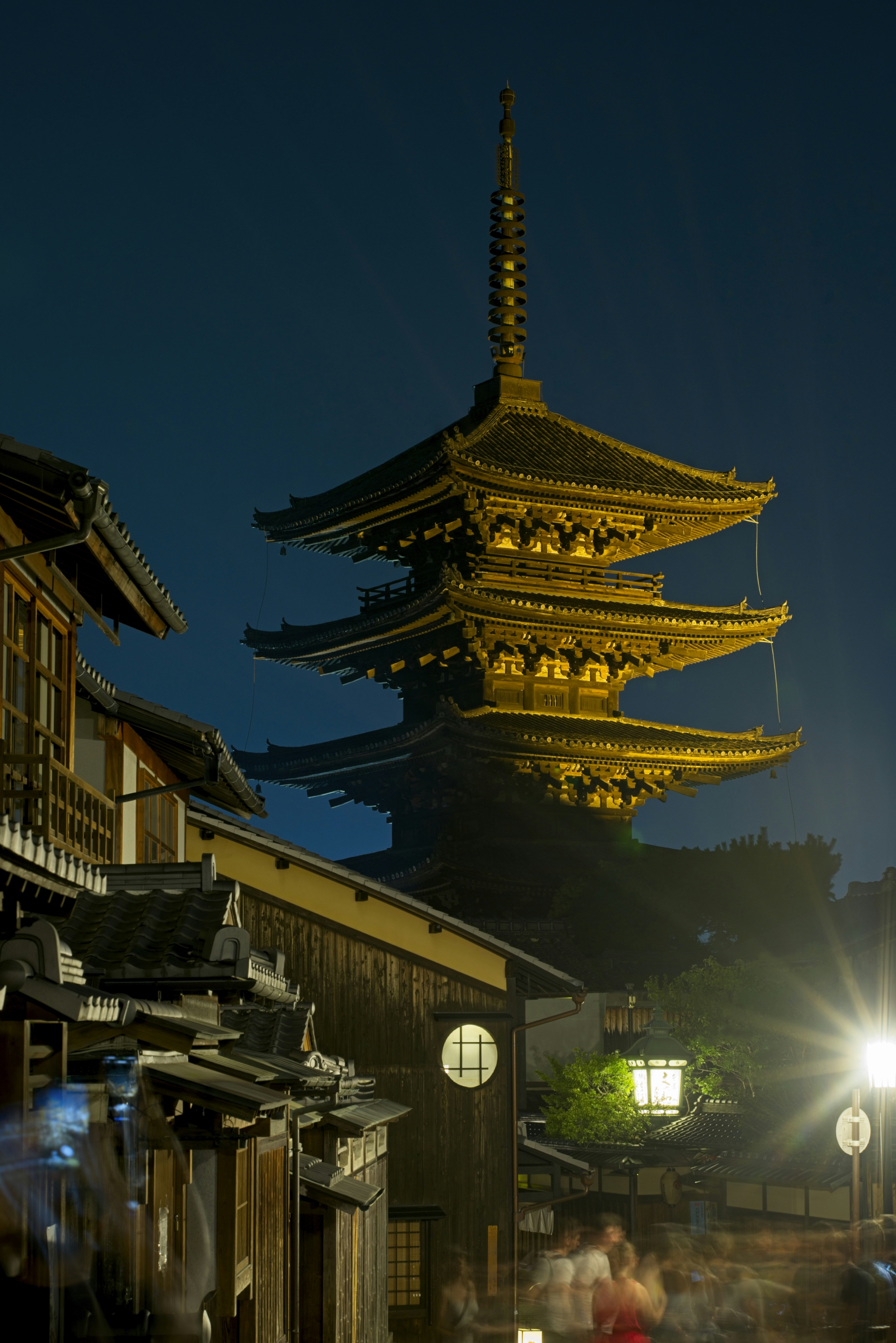 A long exposure captures the illuminated five-story Yasaka Pagoda of Hōkan-ji Temple rising above traditional wooden machiya rooftops in Kyoto's historic Higashiyama district on a humid July evening. Golden light bathes the intricate tiered eaves and ornate finial against the deep blue sky, while streaking light trails and blurred figures below reveal the steady flow of pedestrians through this timeless Kyoto street after dark.