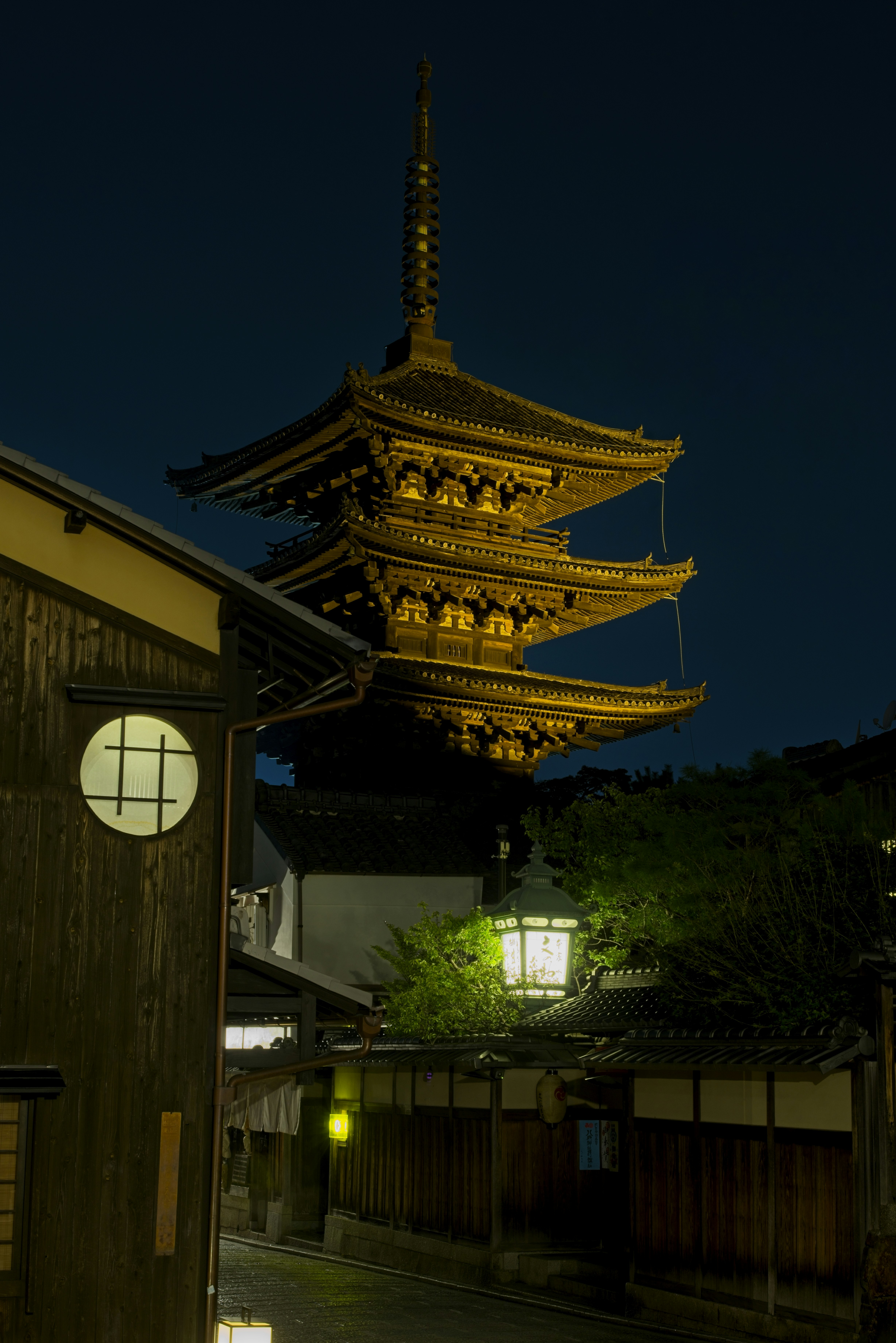 The illuminated five-story Yasaka Pagoda of Hōkan-ji Temple rises against a deep blue night sky, its ornate tiered roofs glowing warmly above the rooftops of Higashiyama's traditional wooden houses. A round latticed window on a nearby machiya facade echoes the pagoda's timeless elegance, while a softly lit stone lantern nestles among trees below. The long exposure smooths the humid July evening into stillness, capturing centuries-old architecture standing quietly over Kyoto's historic streets after dark.