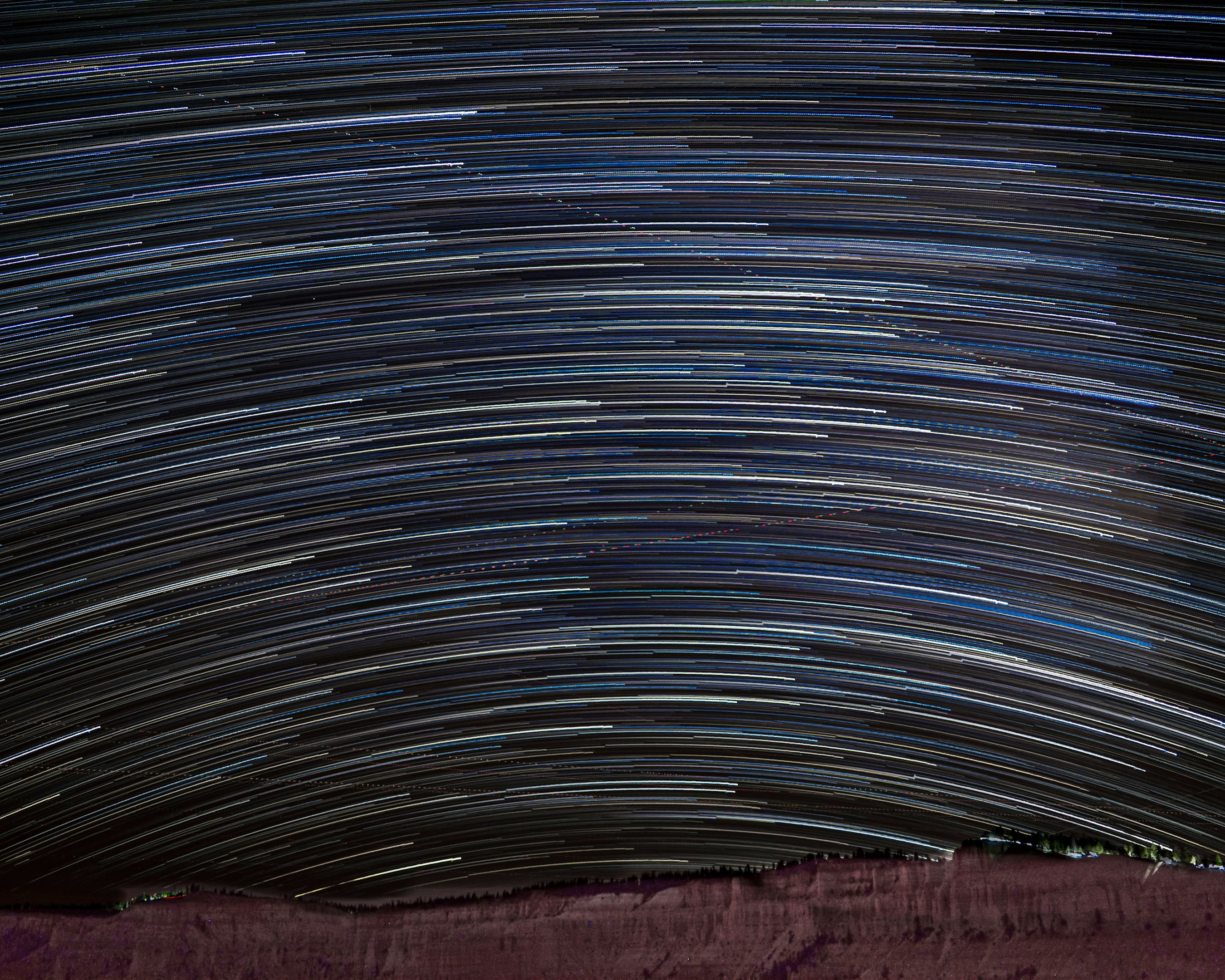 Long-exposure star trails arc across the night sky above a desert ridge, revealing continuous stellar movement and a hint of ground texture at the horizon.
