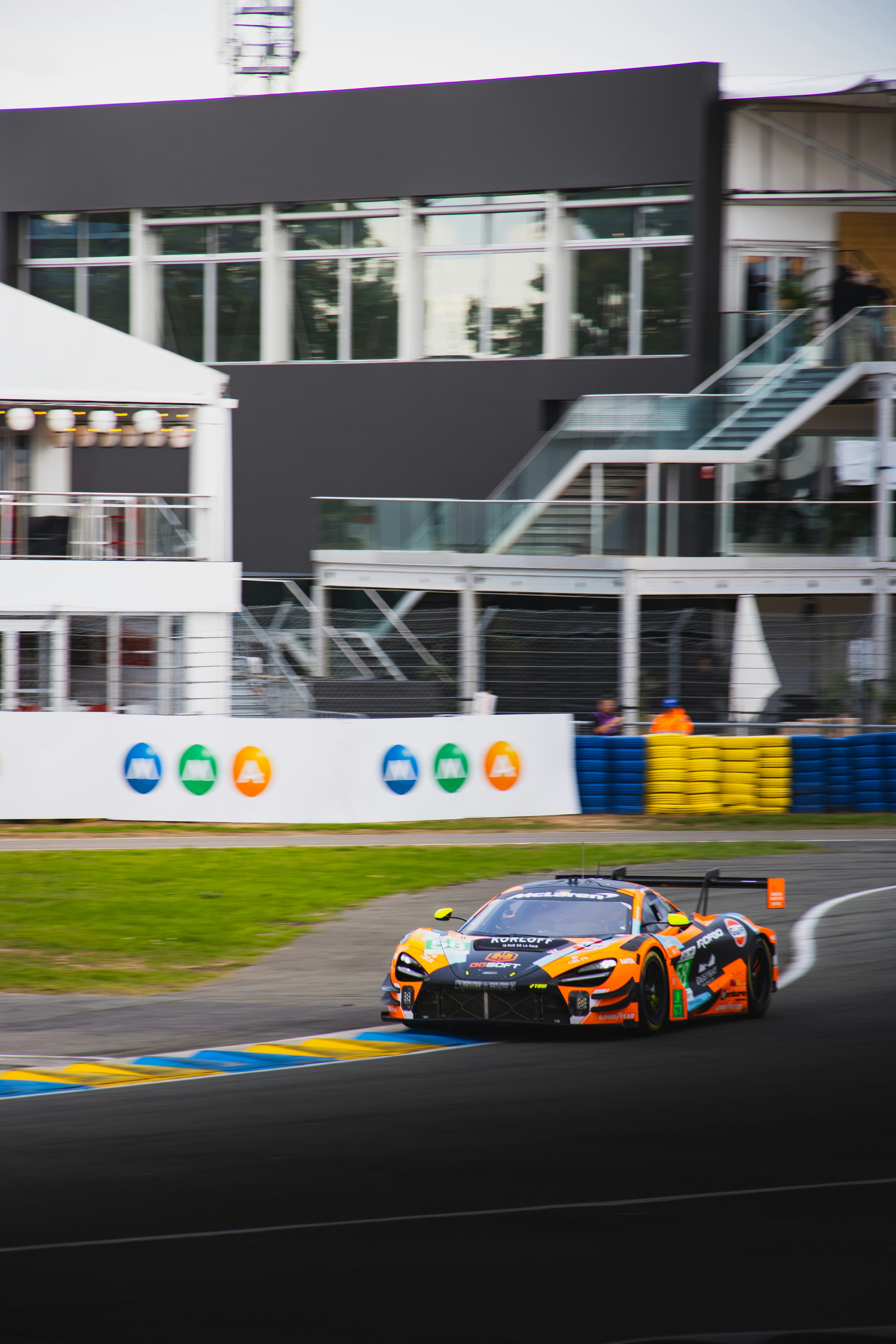 Orange racecar speeds along a track with modern pit building and colorful barrier art in the background.