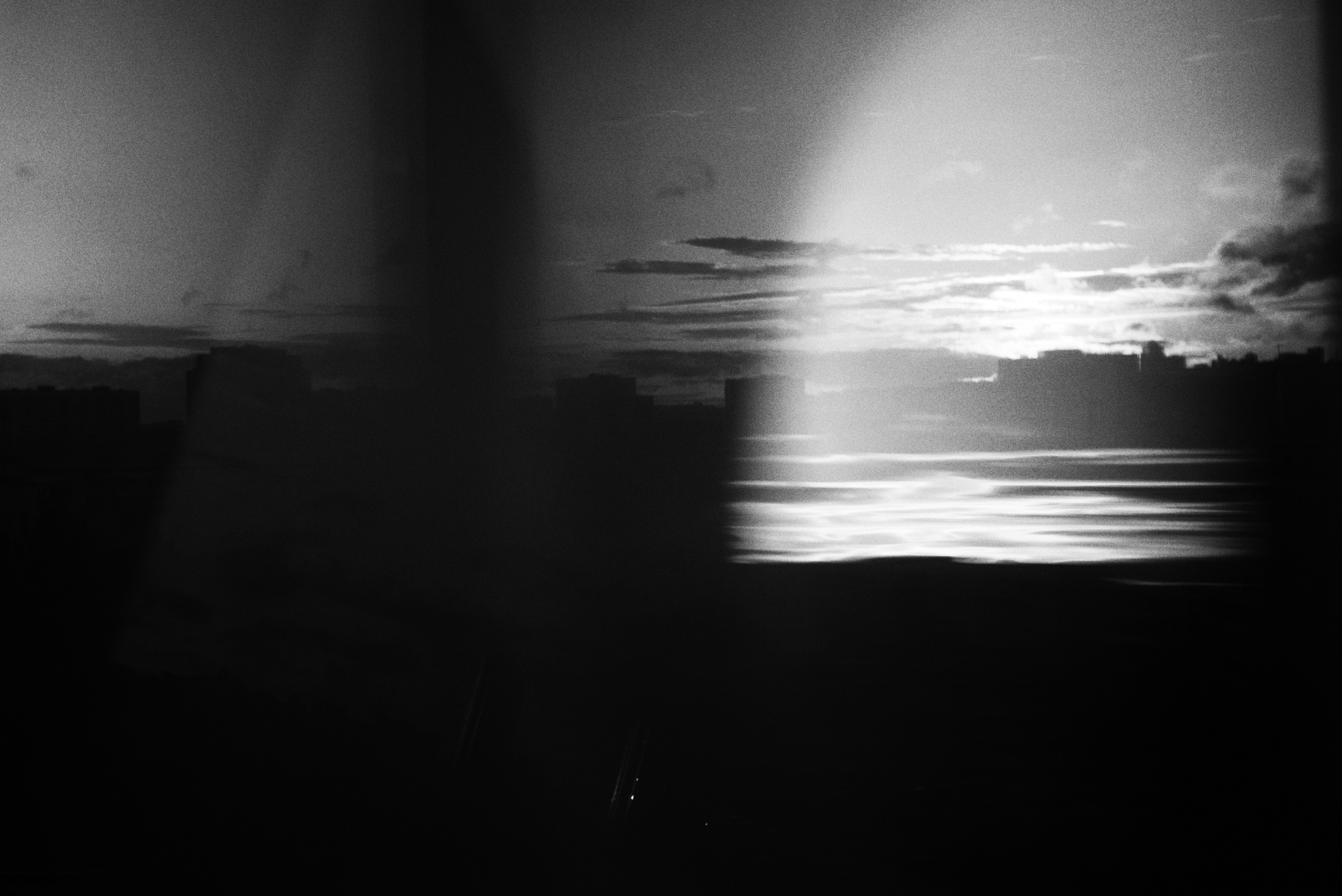 Monochrome horizon over a calm body of water with soft cloud silhouettes; a moody, minimal scene emphasizing silhouette and light play.