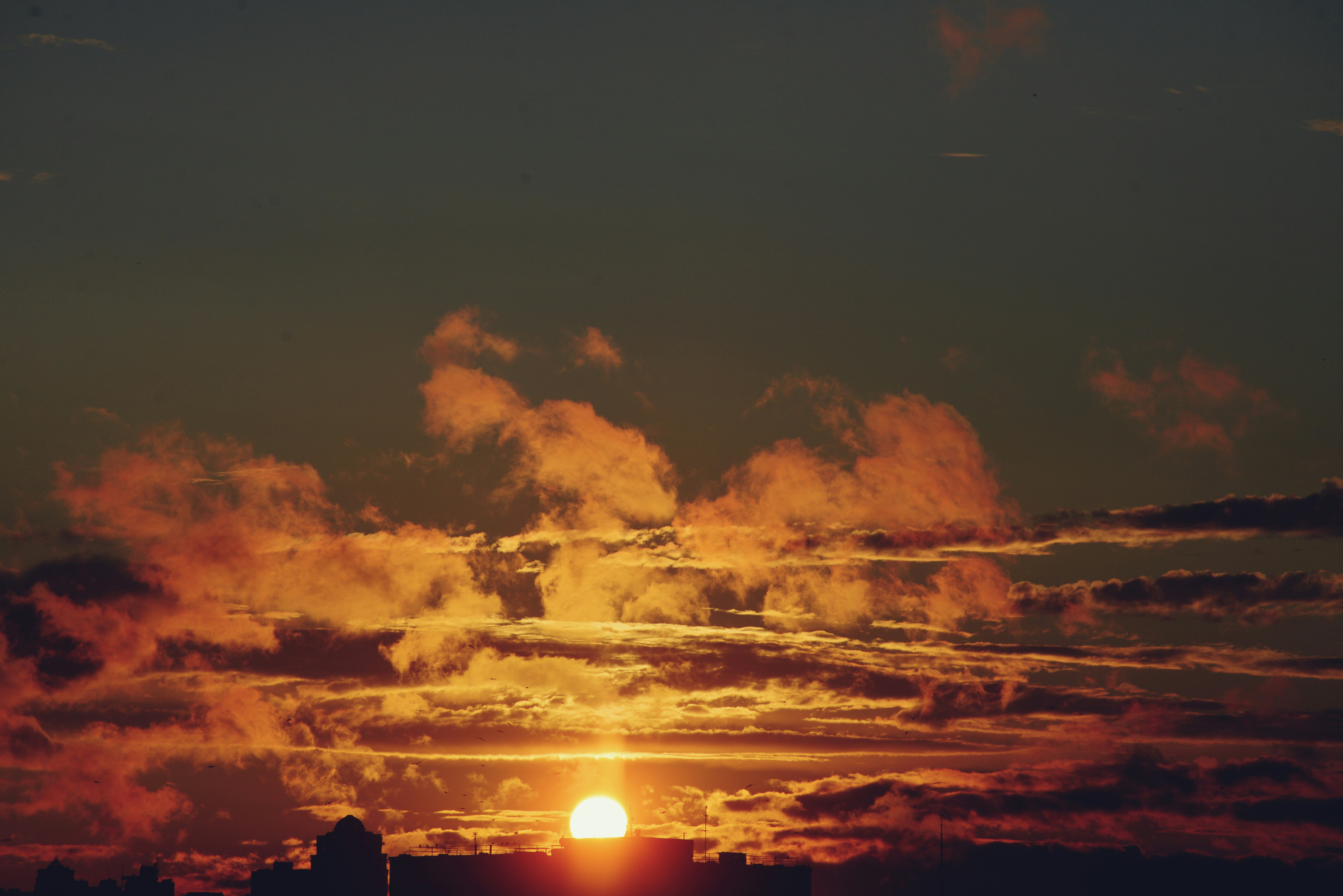 Sun setting over a distant skyline with a bright sun dipping below the horizon; warm orange clouds fill the sky, creating a tranquil city silhouette.