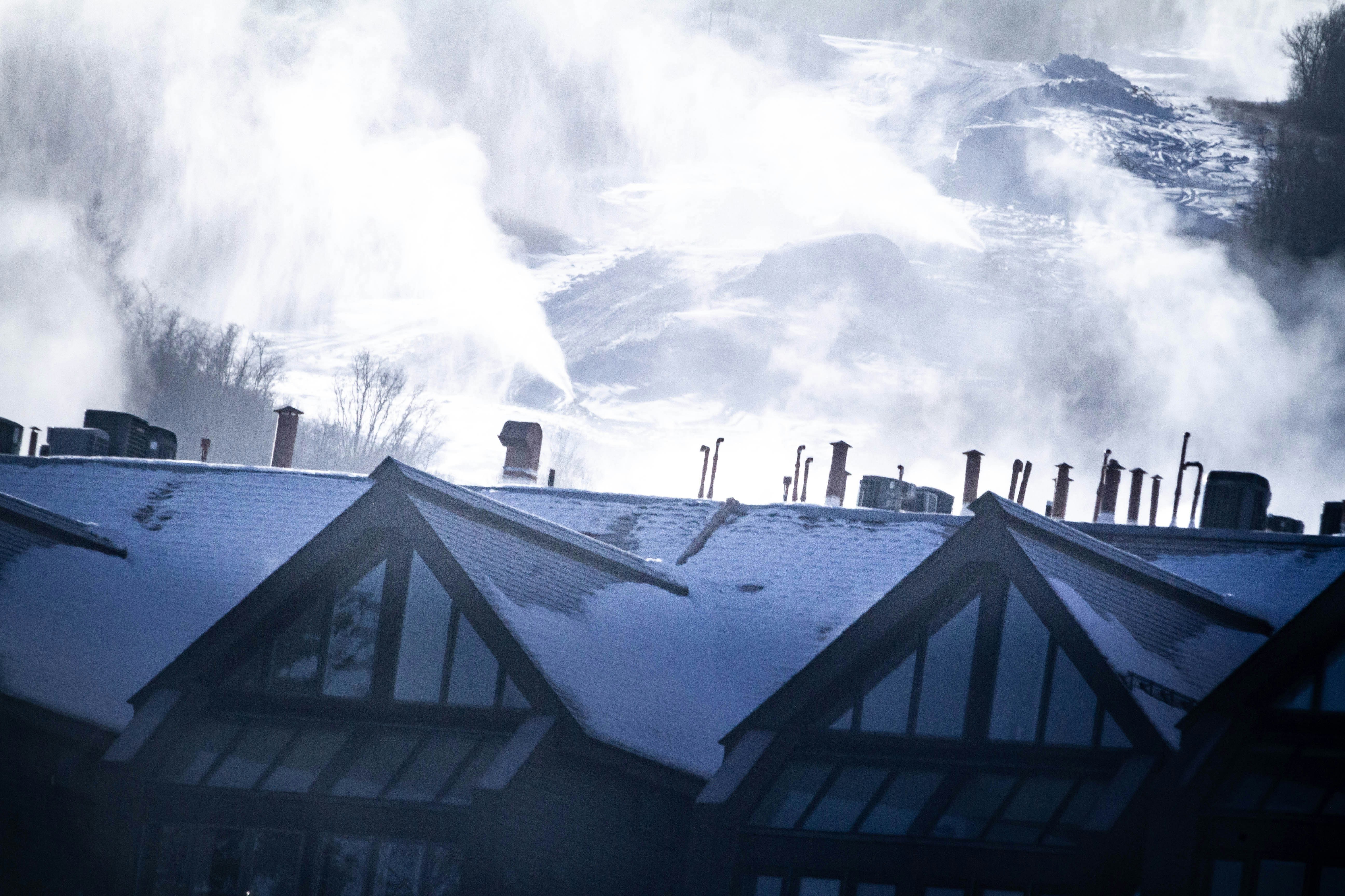 Snow-capped rooftops meet a dramatic plume of steam or mist rising behind a row of chalet-style houses, hinting at a cold urban scene with warm interiors.