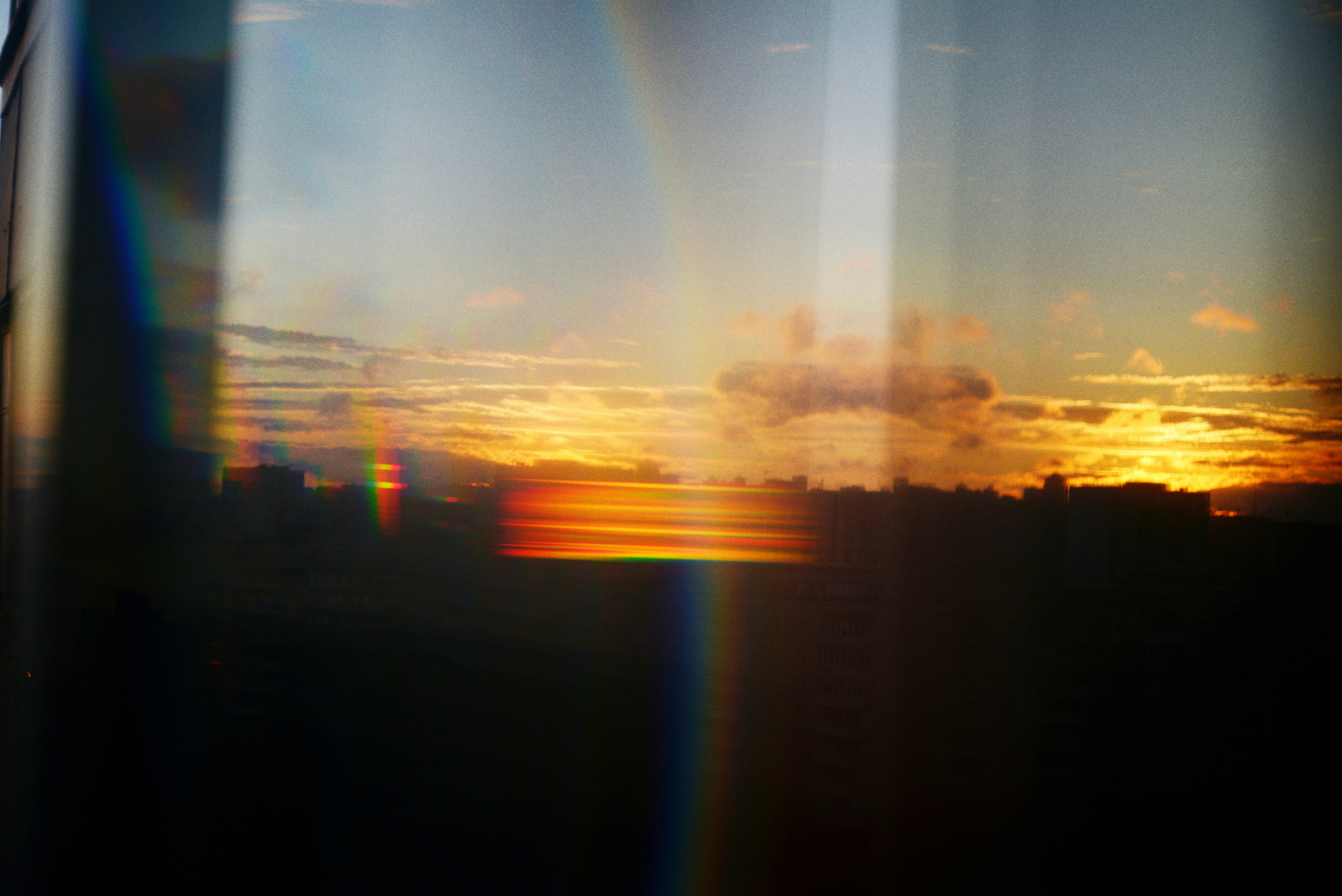 Soft sunset glow with city silhouette and color fringes seen through a glass surface; abstract landscape with blown highlights and vertical light streaks. This is a photographic capture, not a painting or illustration.