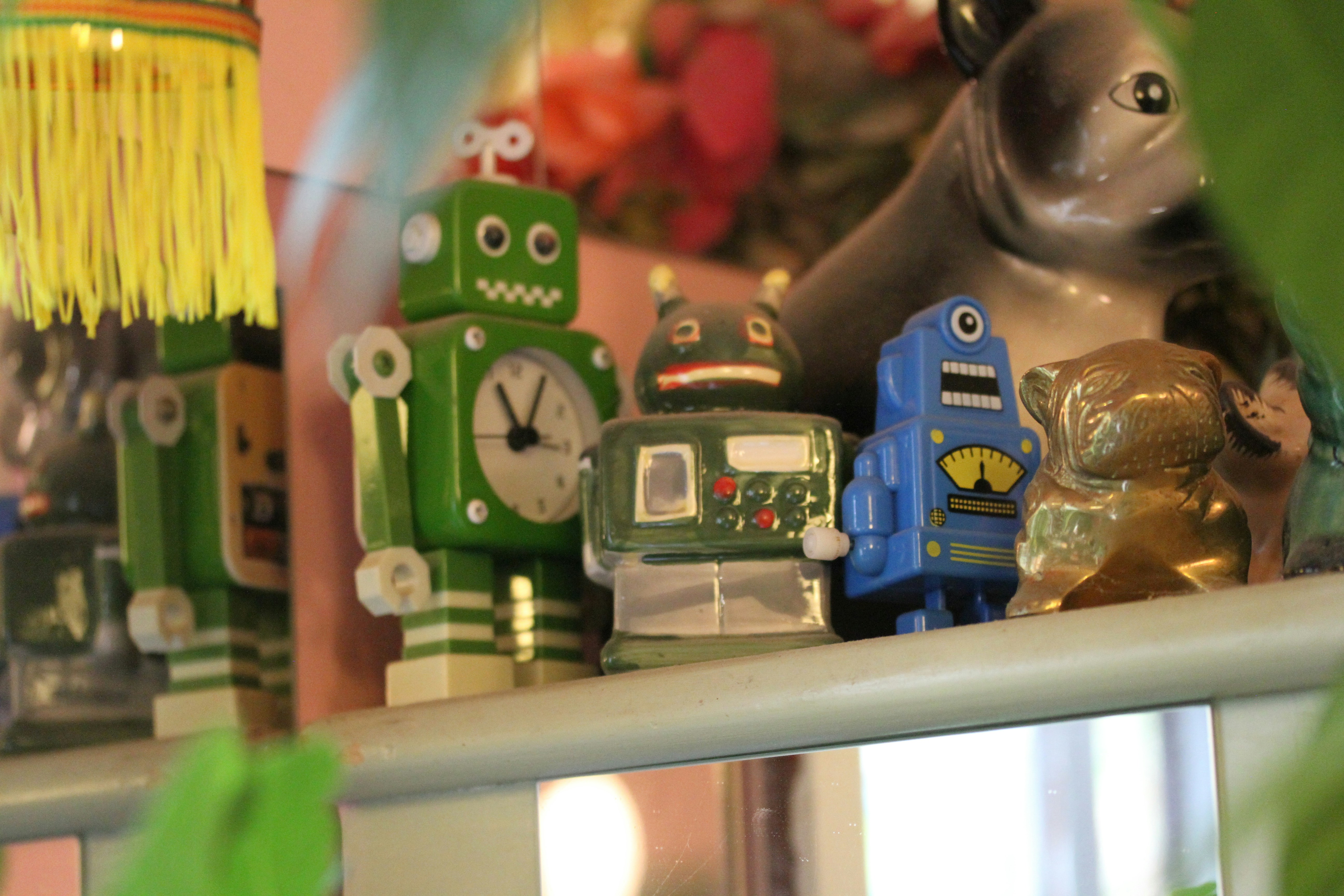 Colorful vintage-style robot figurines lined up on a shelf, with soft bokeh and green leaves in the foreground.