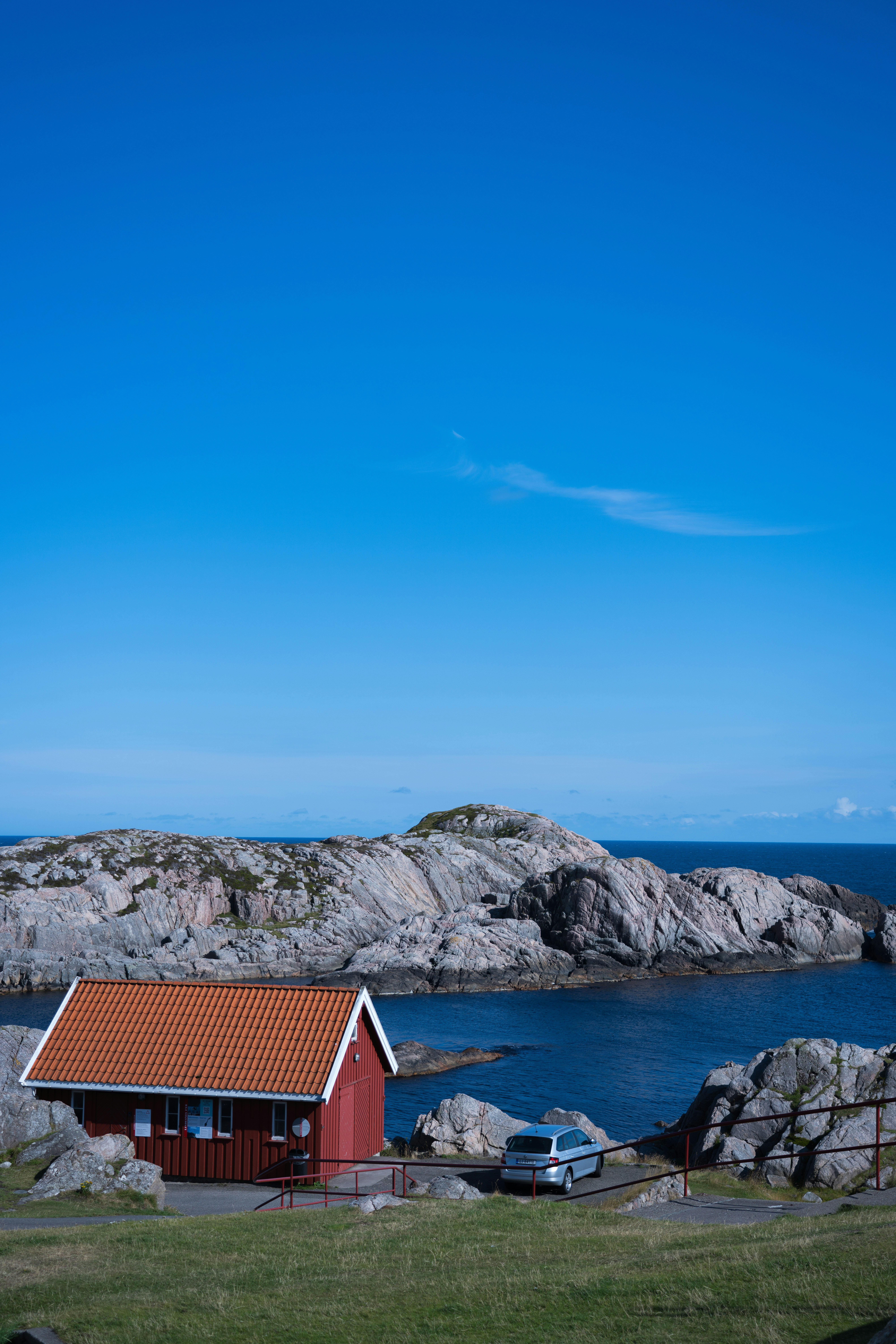 Coastal scene with a small red cabin and parked car set against rugged rocks and calm blue water.