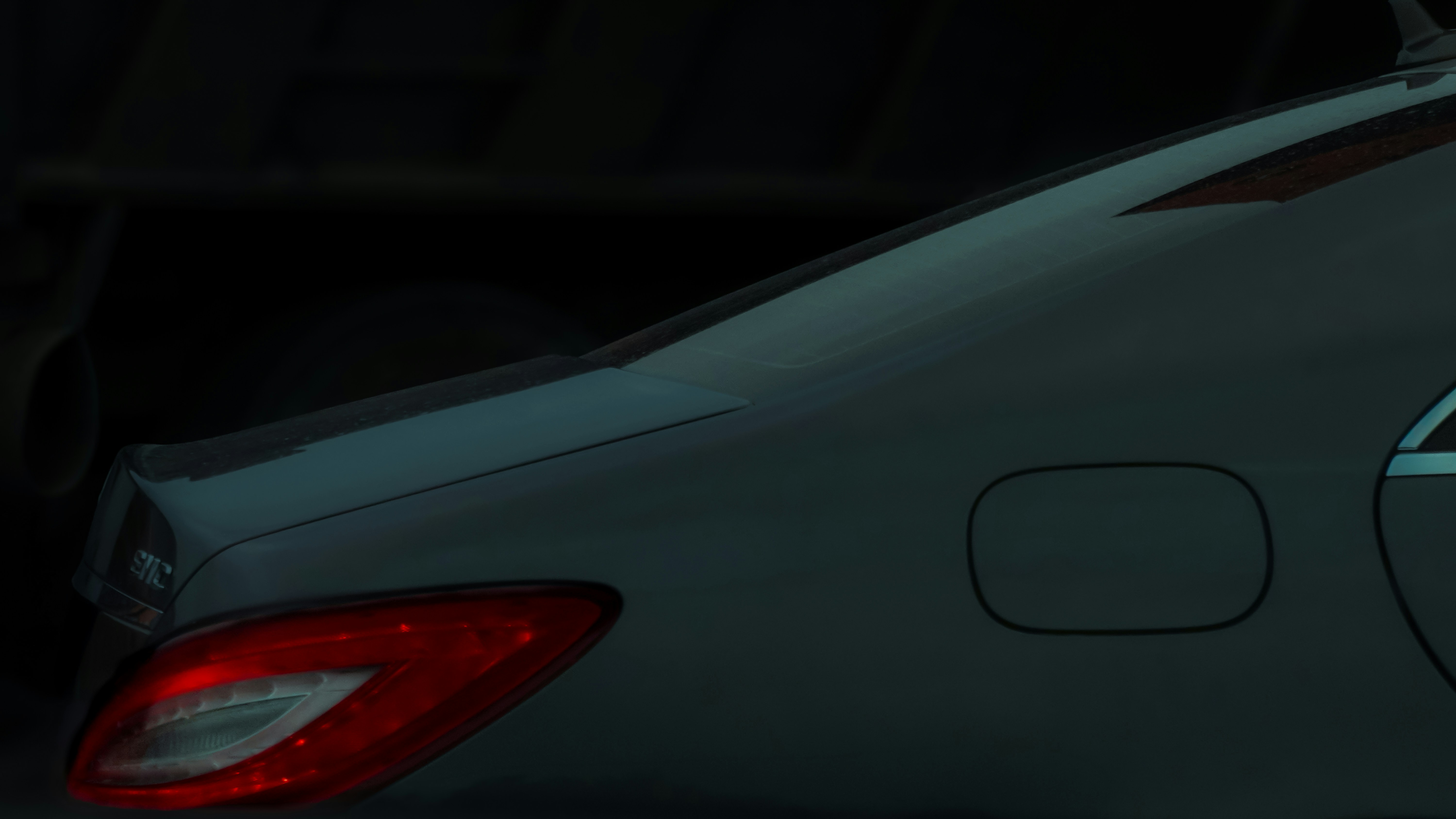 A dark, close-up view of a car's rear section with a red tail light illuminated in low light, showing smooth body contours and a partially visible fuel door.