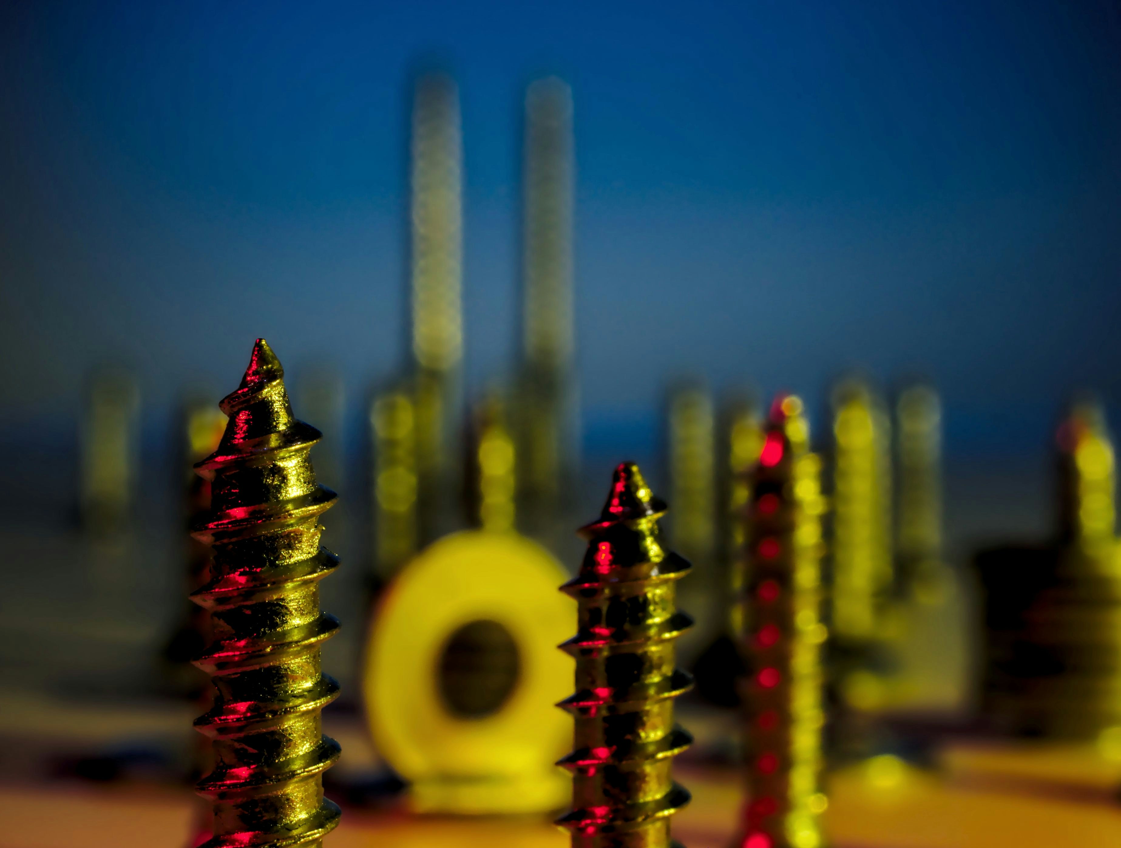Close-up of metallic screws in the foreground with a blurred industrial background, emphasizing texture and color. This appears to be a macro or close-range shot of hardware on a workshop surface.