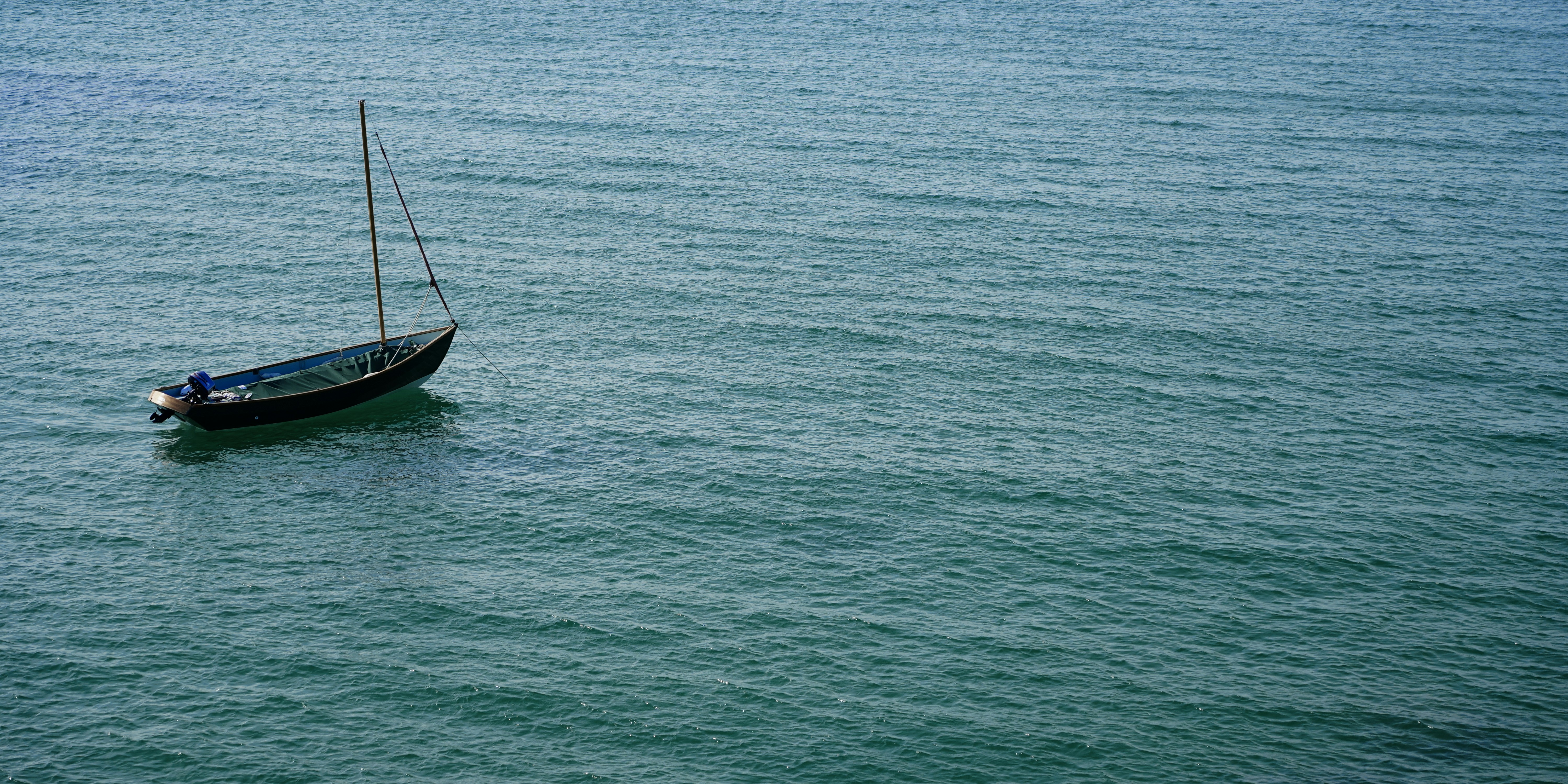 A solitary boat sits on calm turquoise water, occupying the upper-left quadrant and creating negative space around it. A minimal seascape emphasizing serenity and simplicity.