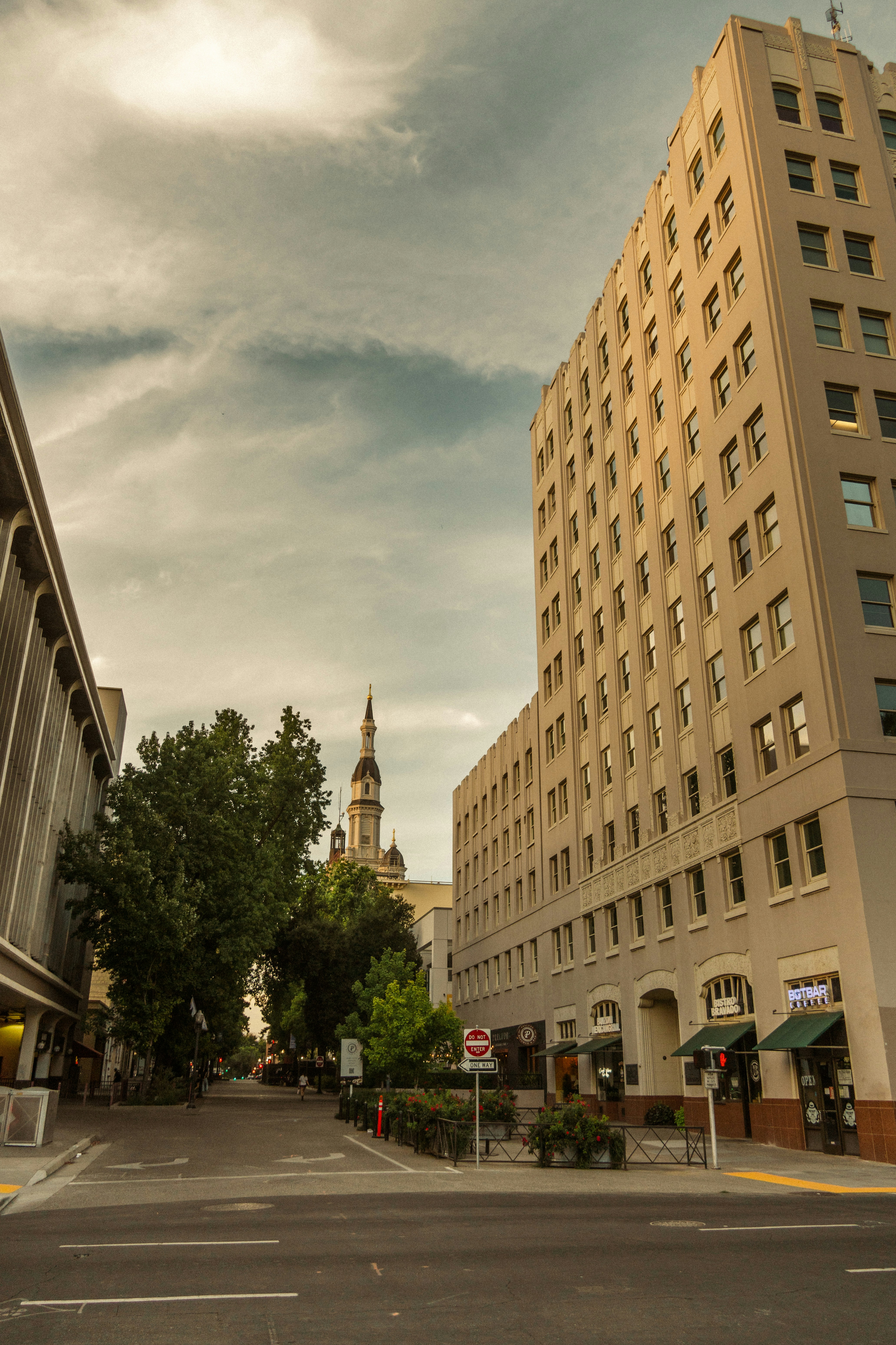 Dusk-lit city street with tall beige buildings flank a tree-lined corridor and a distant clock tower, conveying a calm urban moment.