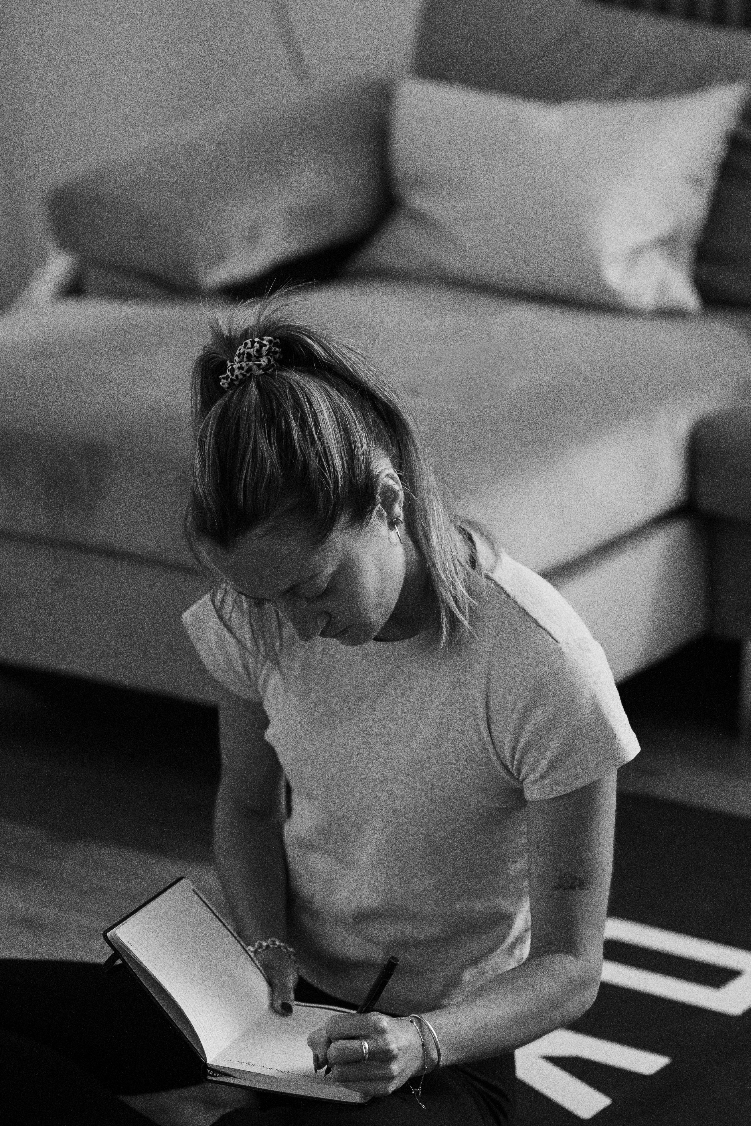 A girl sits on the floor, writing in an open notebook with a pencil, in a grayscale interior setting with a sofa in the background.