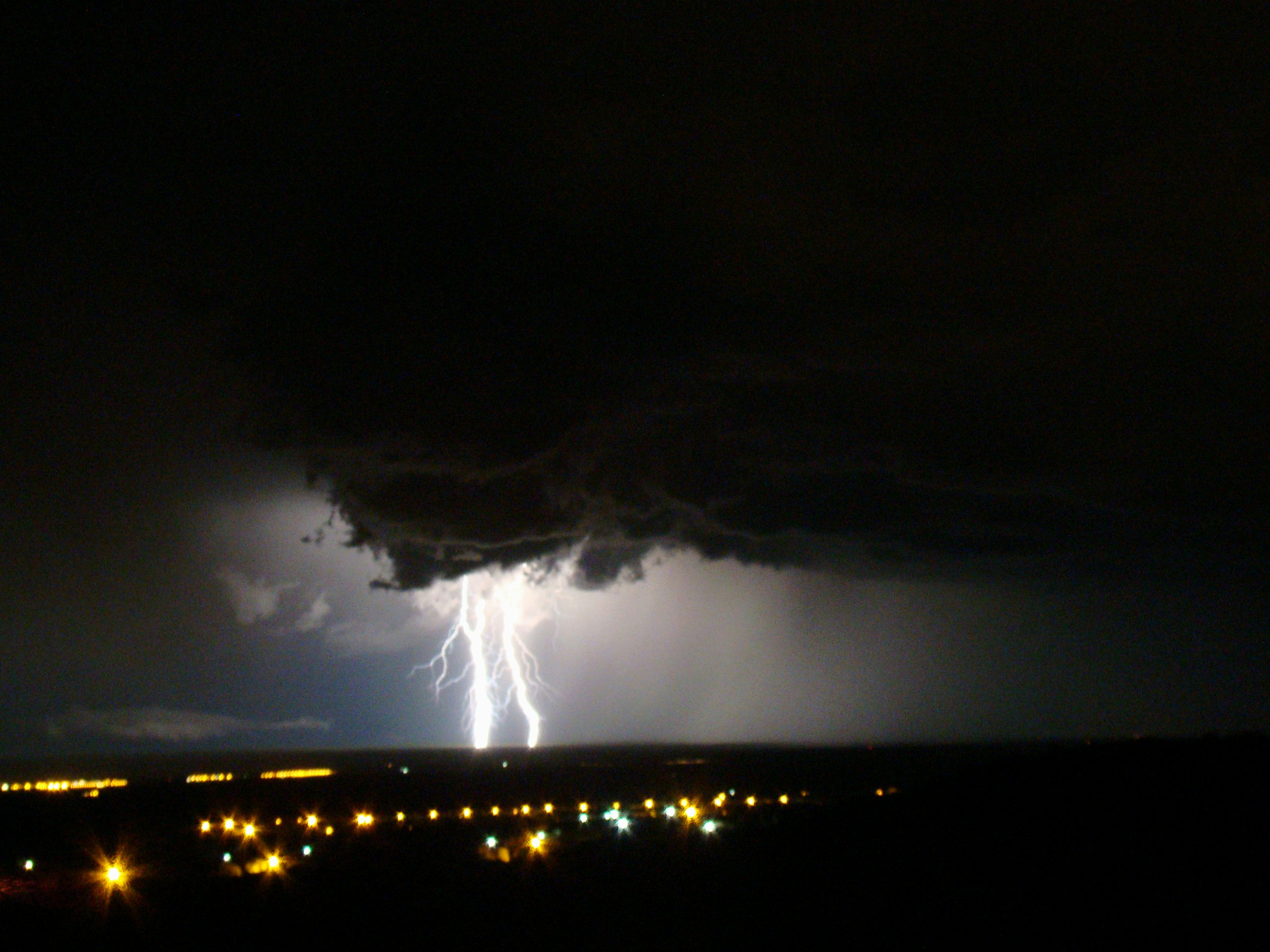 Lightning bolt strikes over a dark horizon with city lights beneath; a stormy sky dominates the scene, highlighting dramatic contrast between illumination and darkness.