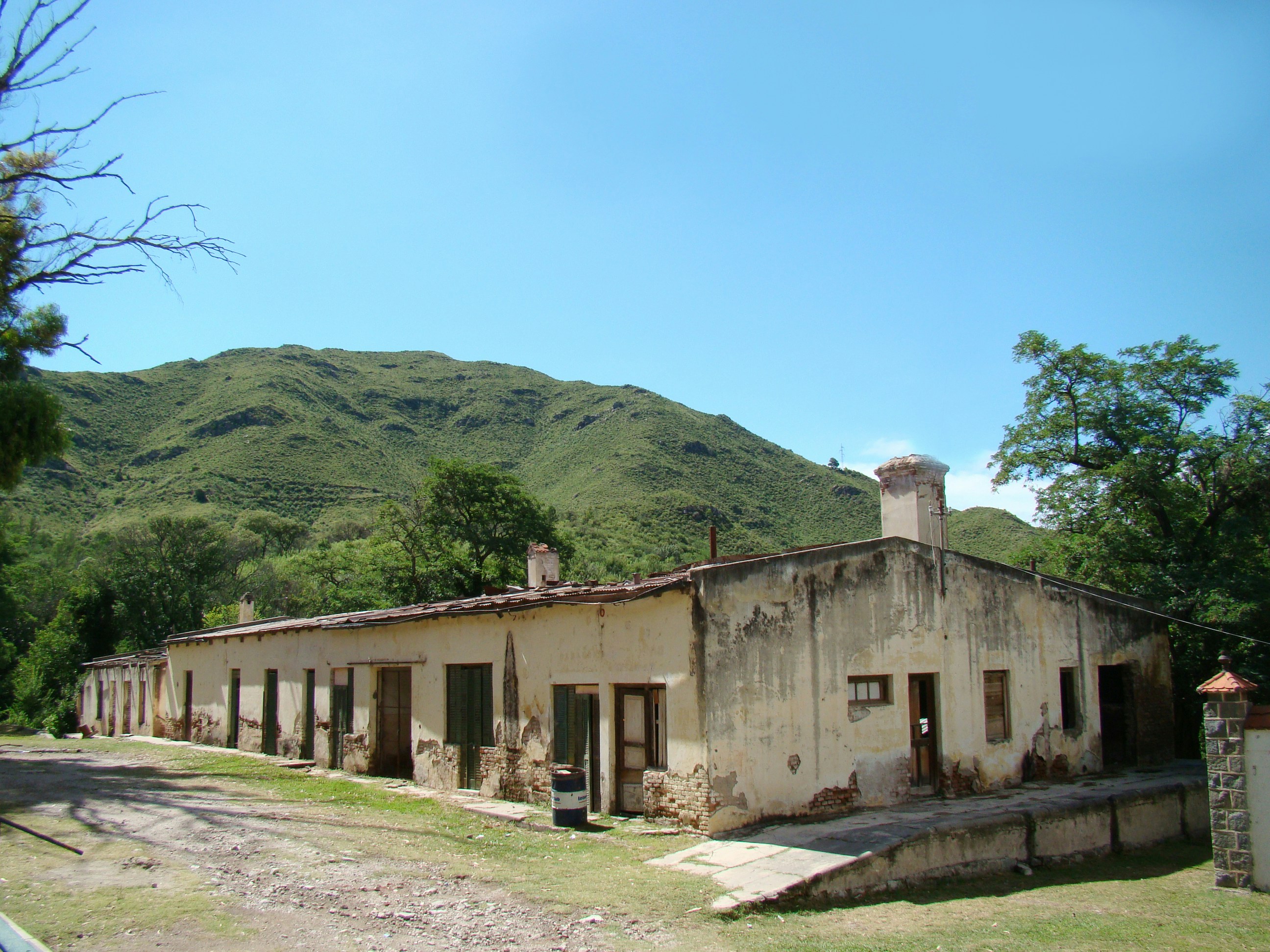 Weathered single-story buildings line a dirt road with a green hillside backdrop and a clear blue sky.