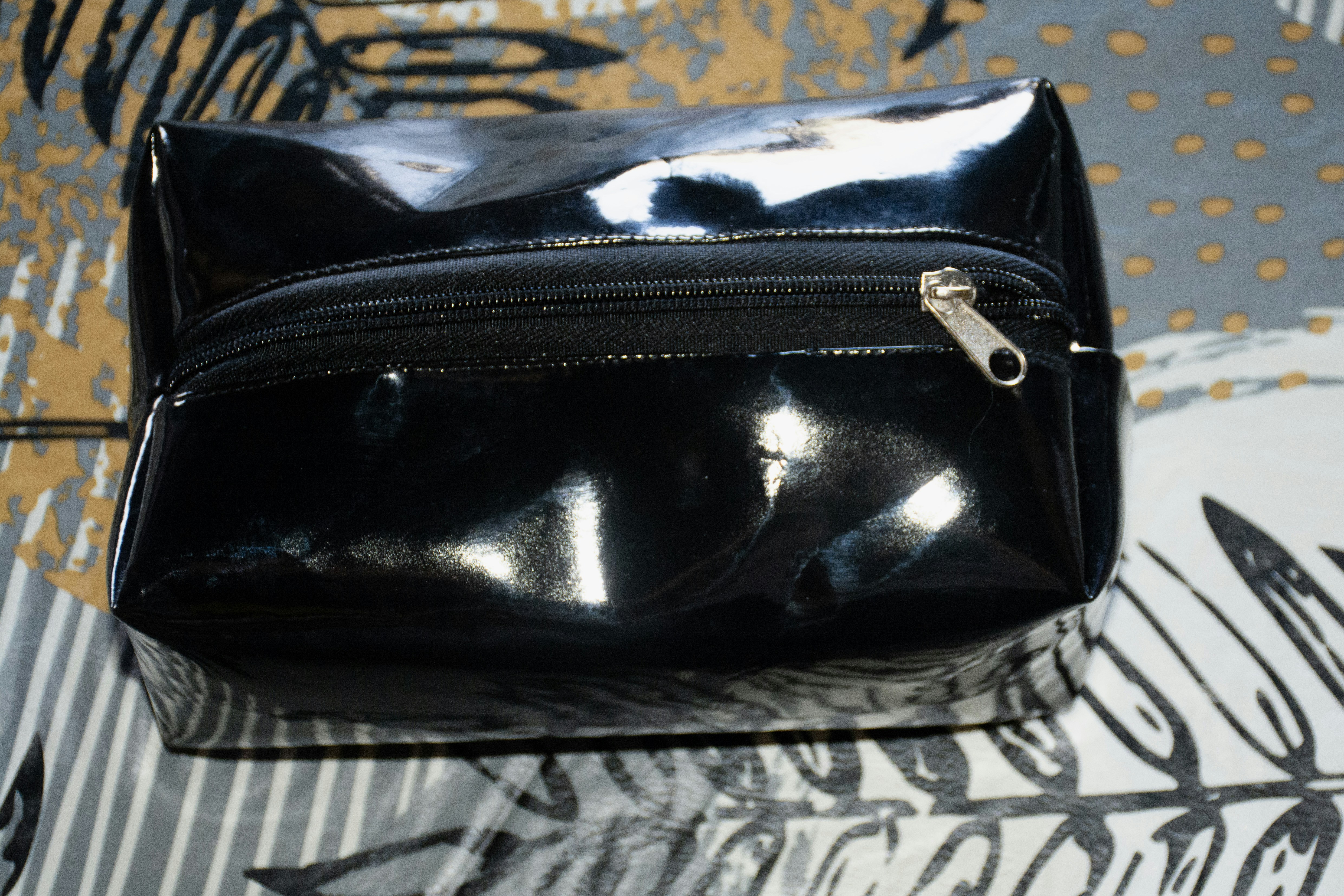 Shiny black toiletry pouch resting on patterned surface with zipper gleam