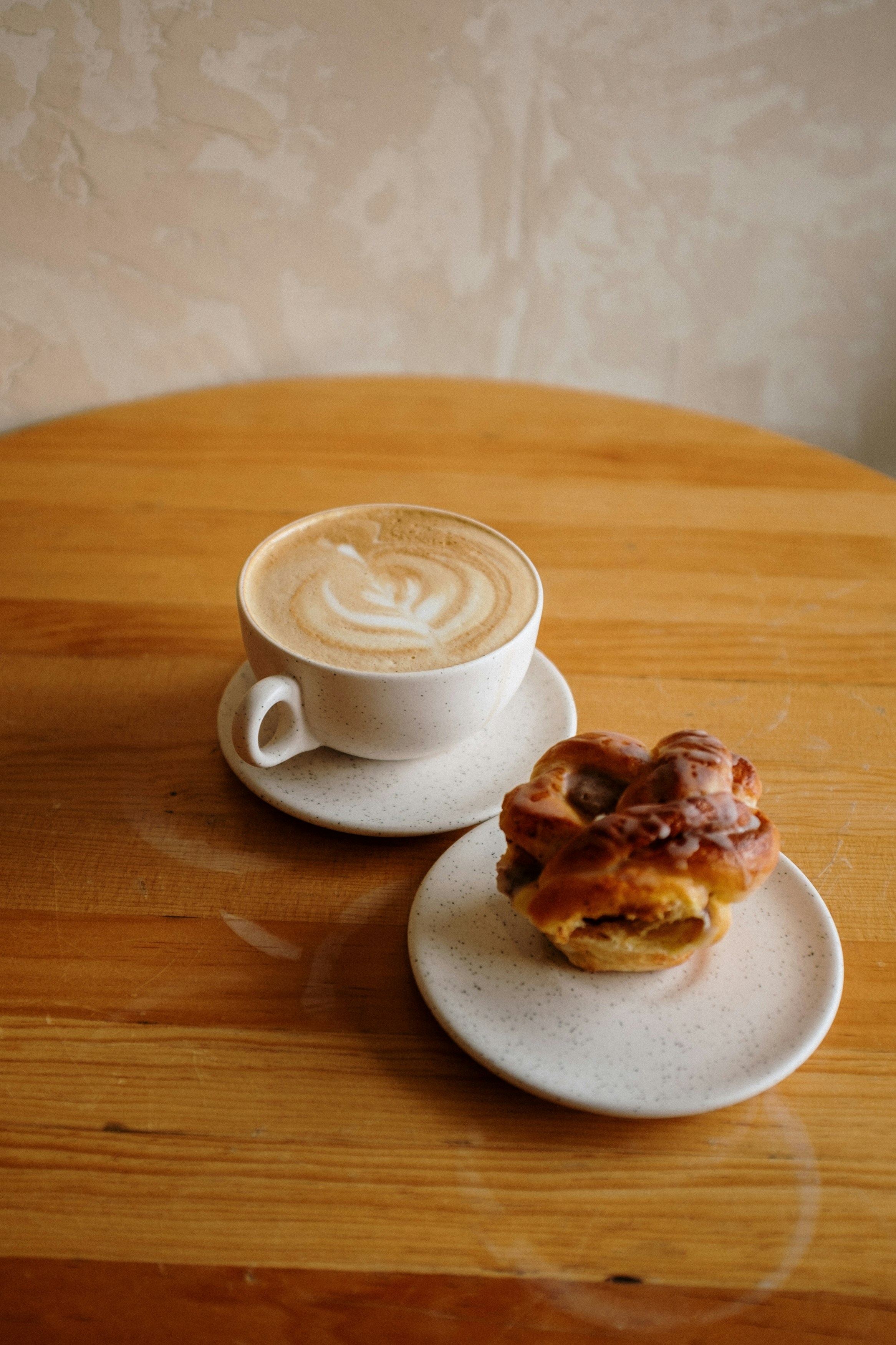 Warm latte and pastry sit on a honey-toned wooden table in a cozy cafe setting. A simple ceramic cup and saucer accompany a small plate with a pastry, suggesting a casual, intimate coffee break.