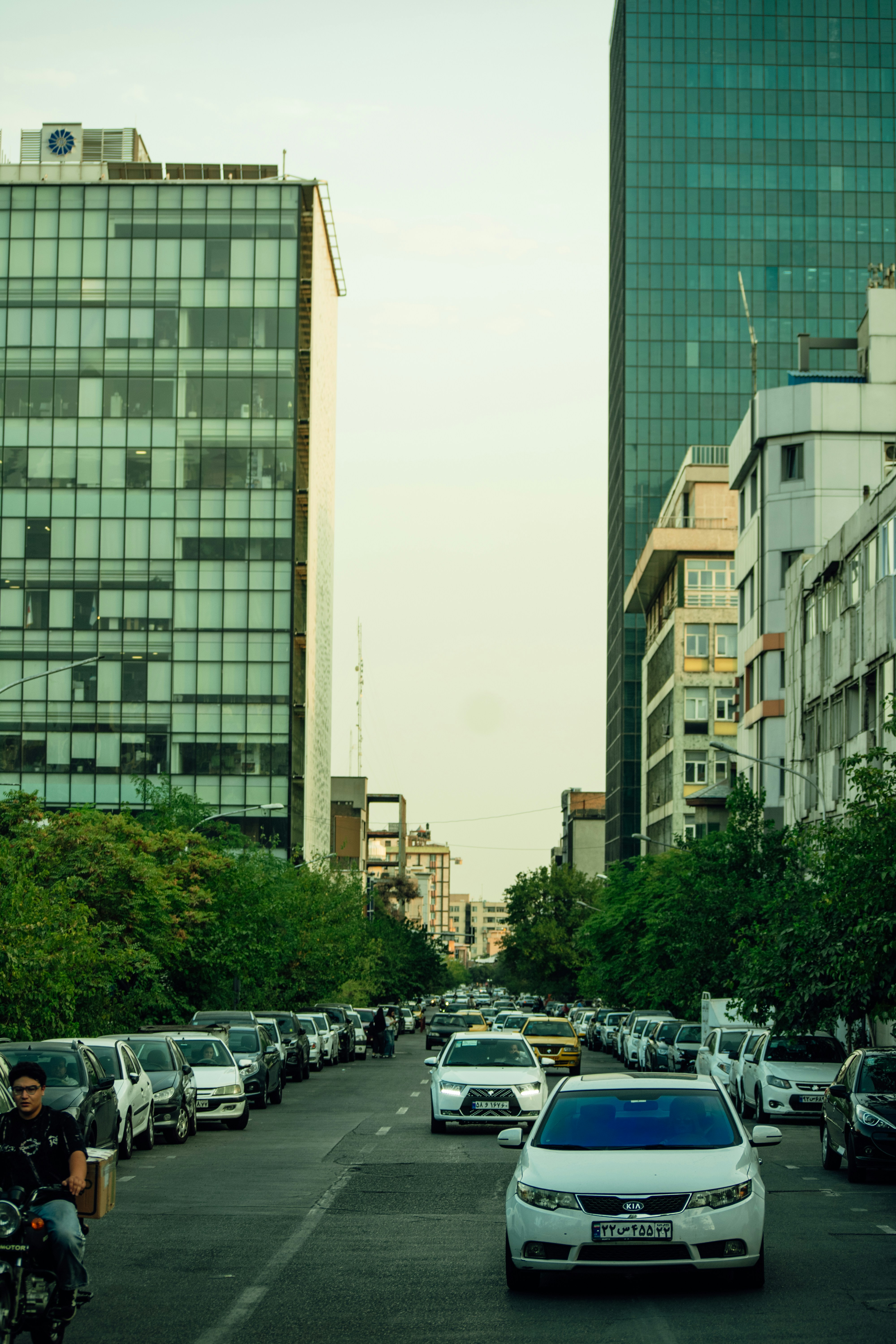 City street lined with parked cars and mixed high-rise buildings, captured in daylight. A sense of depth leads toward a distant vanishing point among the urban canyon.