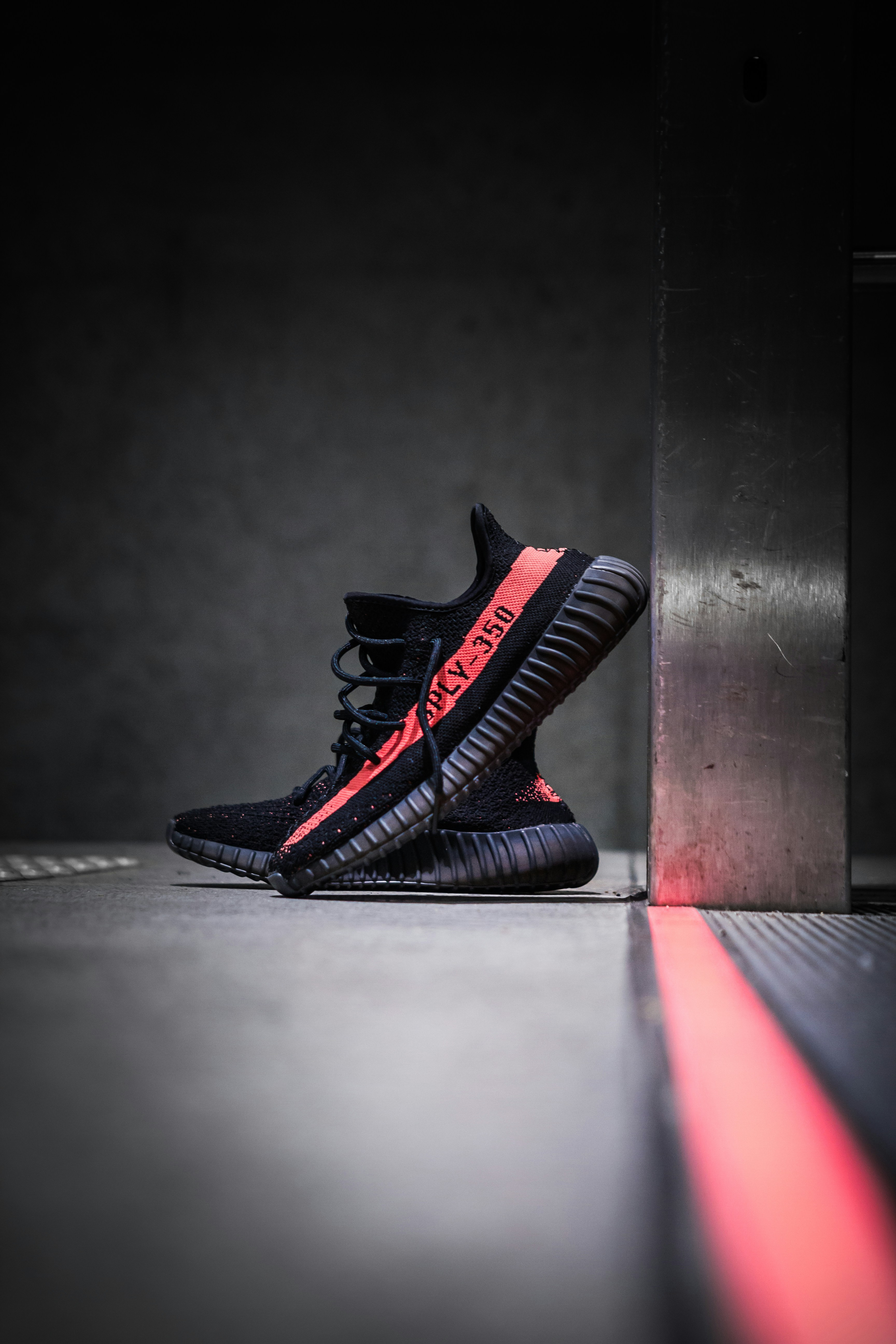 Black sneaker with red stripe sits near a metallic column on a concrete surface, bathed in moody light; a subtle red line guides the eye along the floor. logo-free composition emphasizing texture and contrast.
