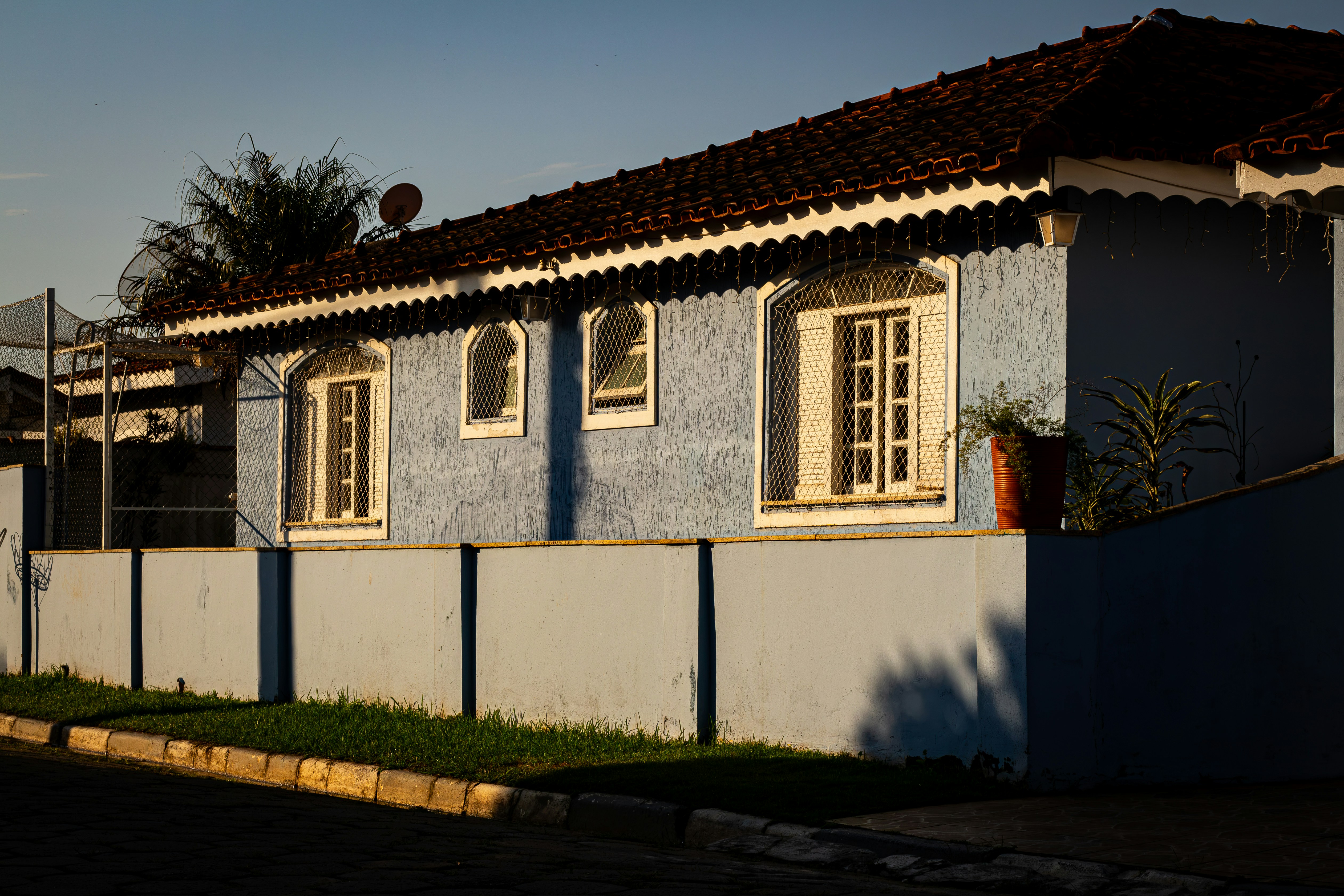 Late afternoon sunlight rakes across the facade of a pale blue house with arched windows, lattice screens and a terracotta tile roof on a quiet cobbled street.