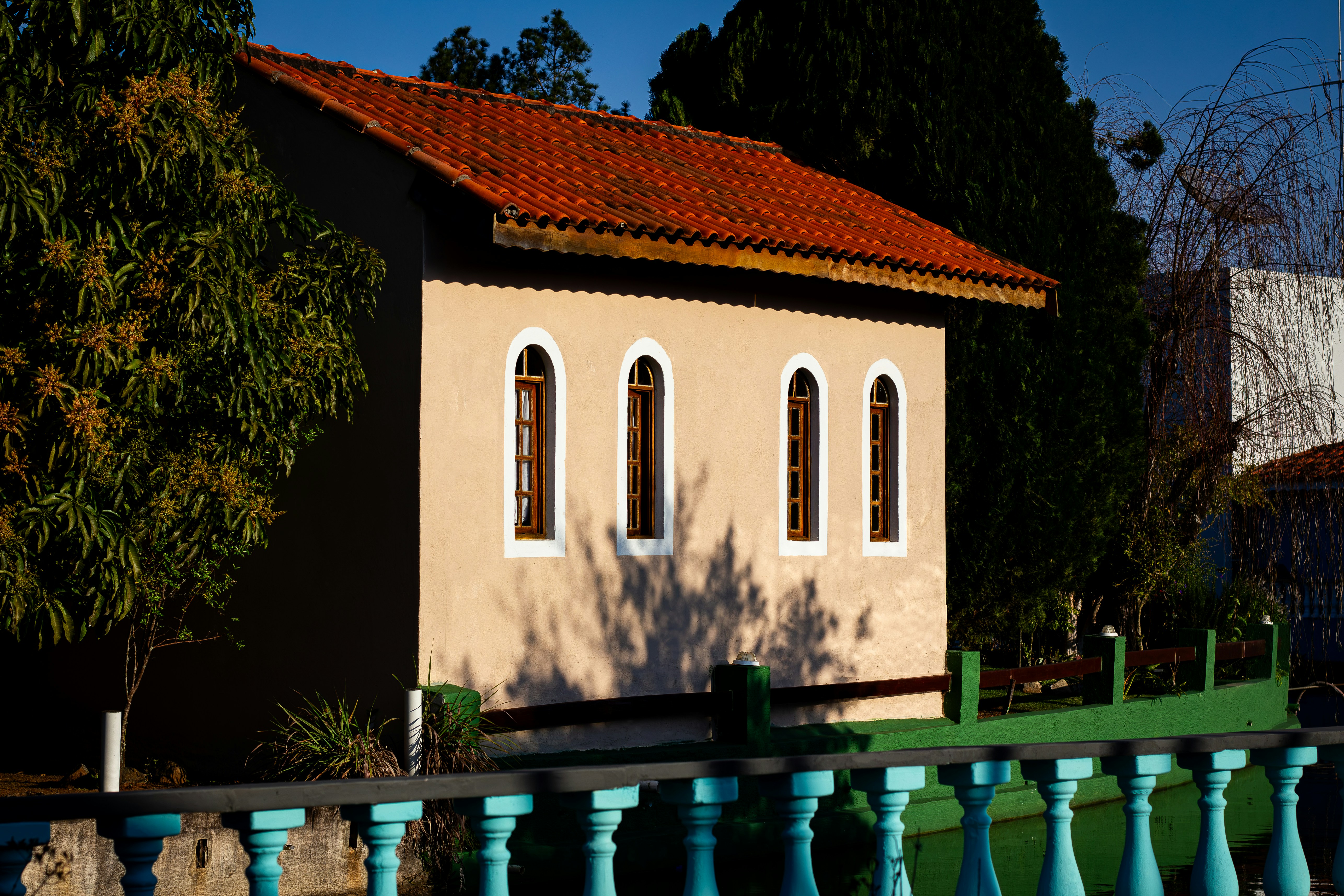 A small building with a red clay tile roof and four narrow arched windows glows in late afternoon light behind a turquoise balustrade, with tree shadows falling across the facade.
