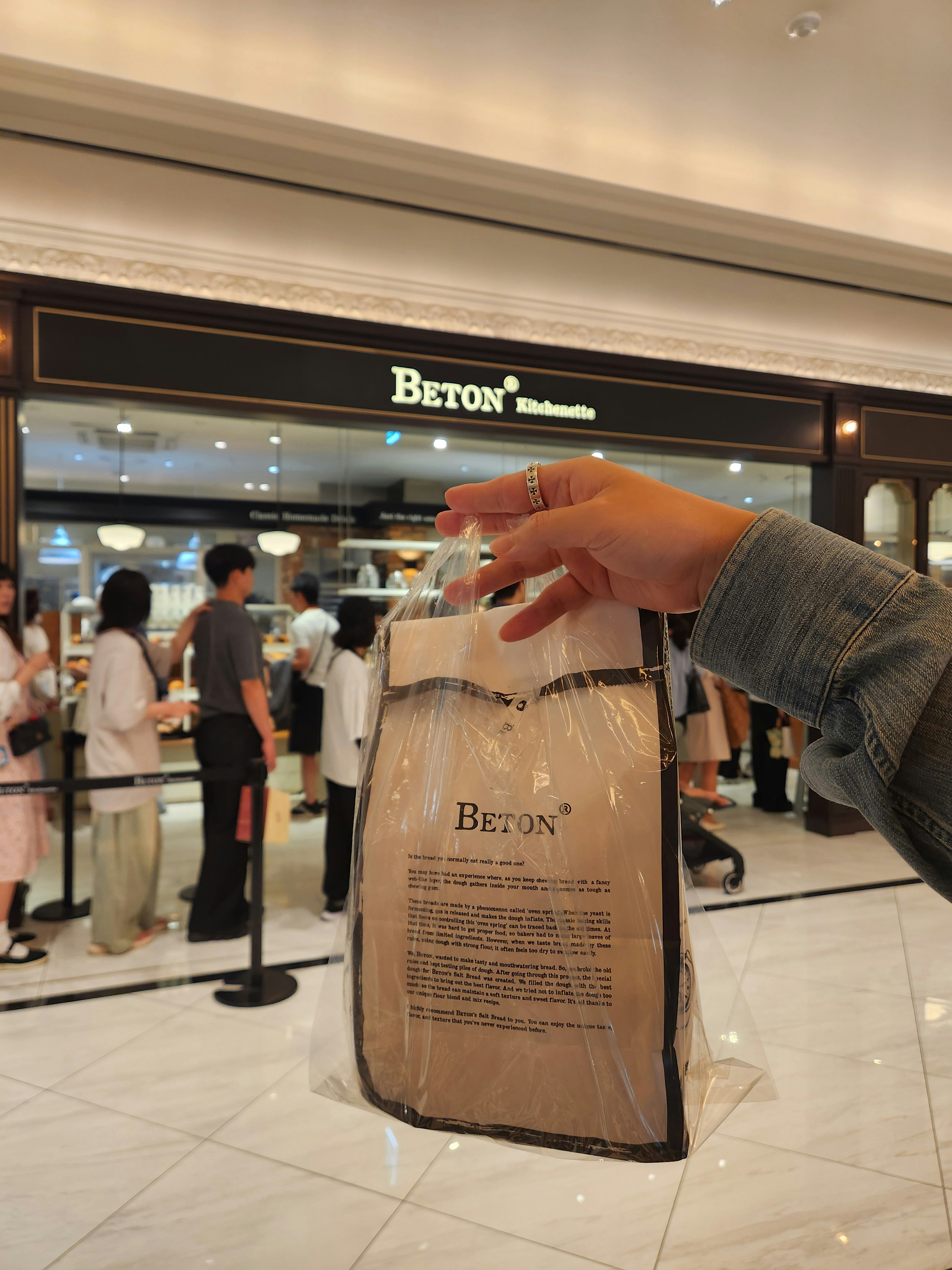 Passport-sized hand-held bag with Beton branding held in foreground inside a shopping mall.