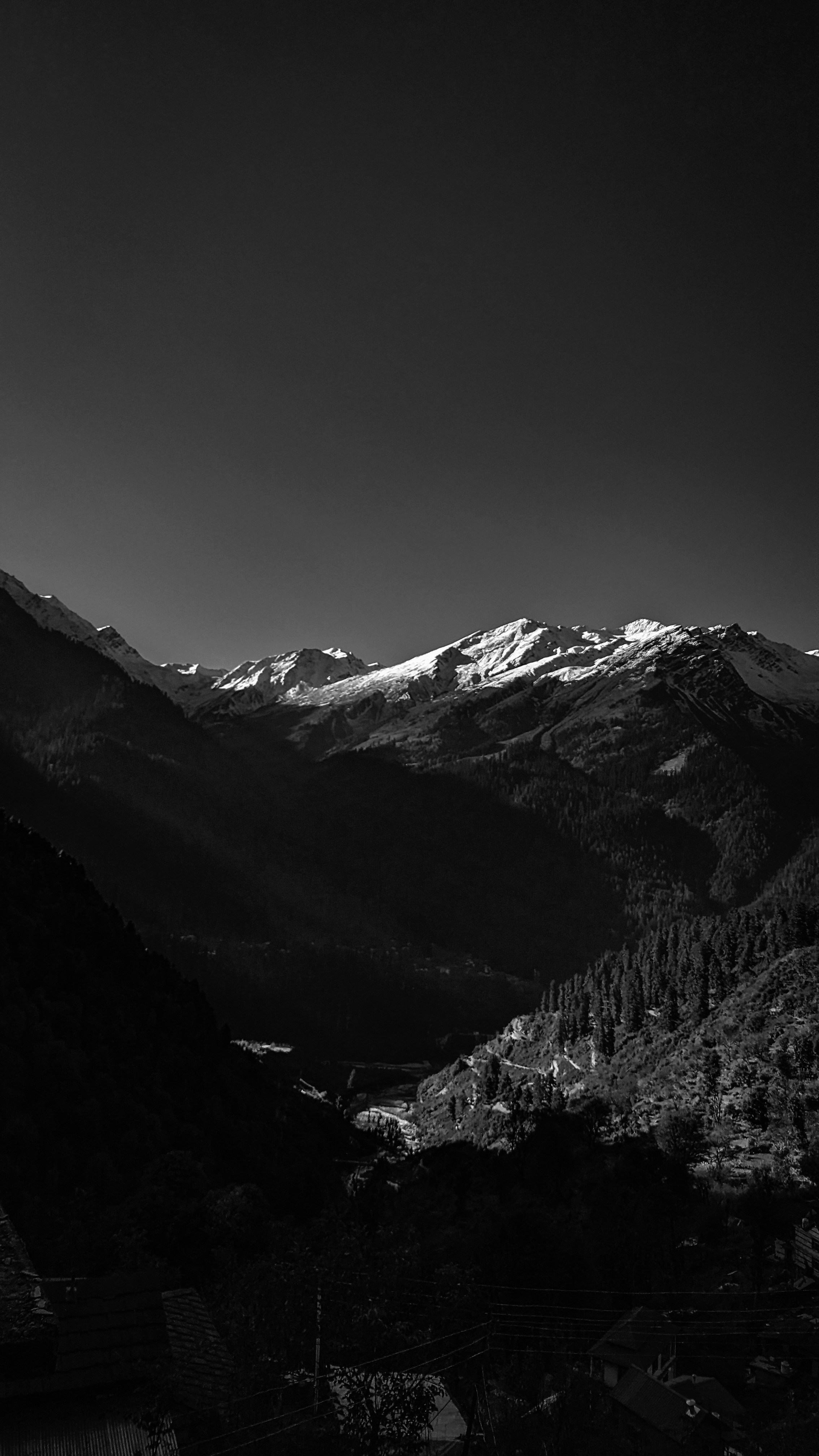 A dramatic black and white view of the mountains surrounding Tosh, Himachal Pradesh. Snow-covered Himalayan peaks rise above the deep forested valley, with strong light and shadow giving the landscape a timeless, cinematic character. Tosh sits in the Parvati Valley and is surrounded by rugged mountains, pine forests and scenic trekking routes, making it a beautiful destination for Himalayan landscape photography and mountain travel.