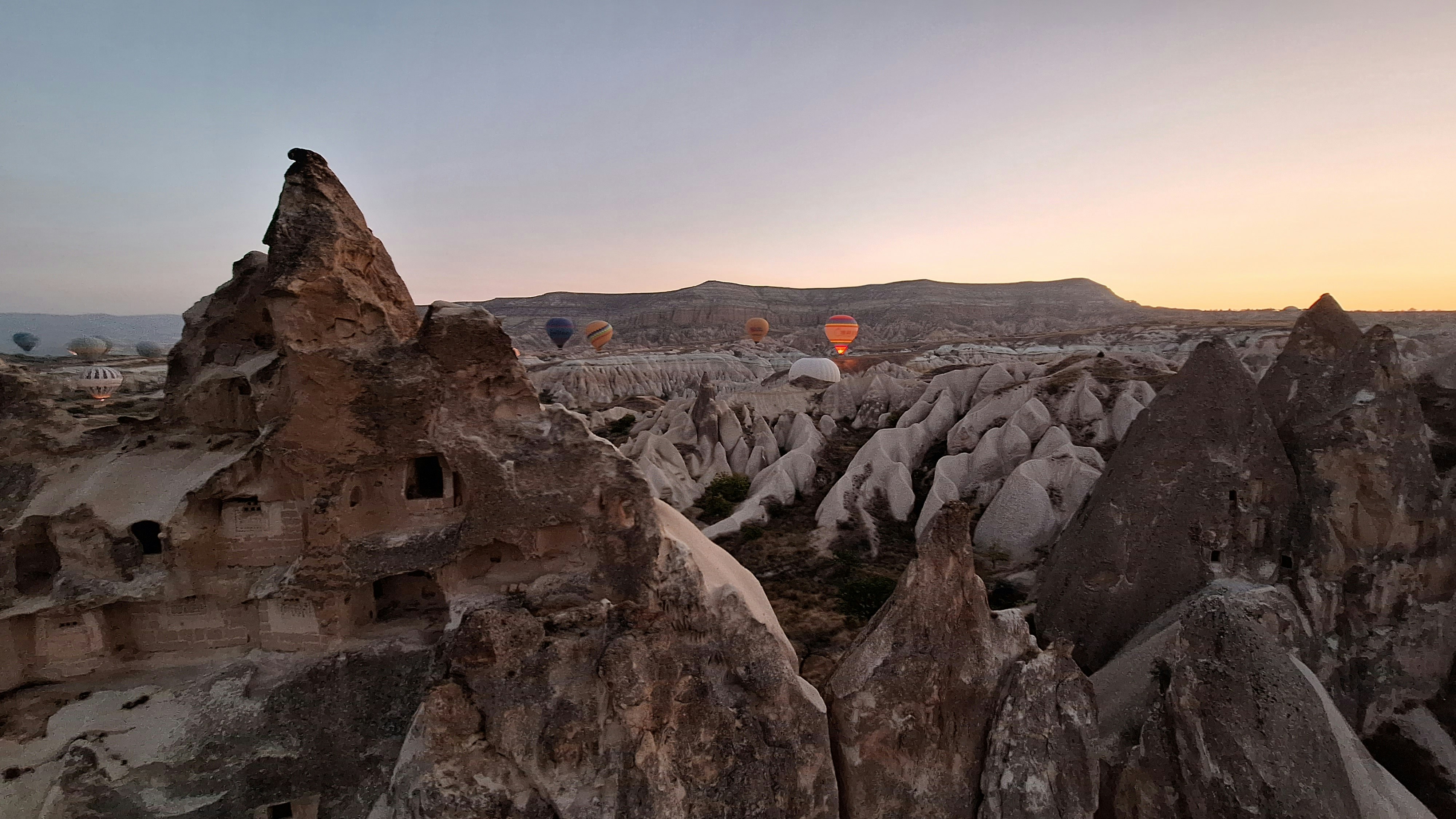 Soft dawn light over carved rock formations and distant balloons rising above the valley.