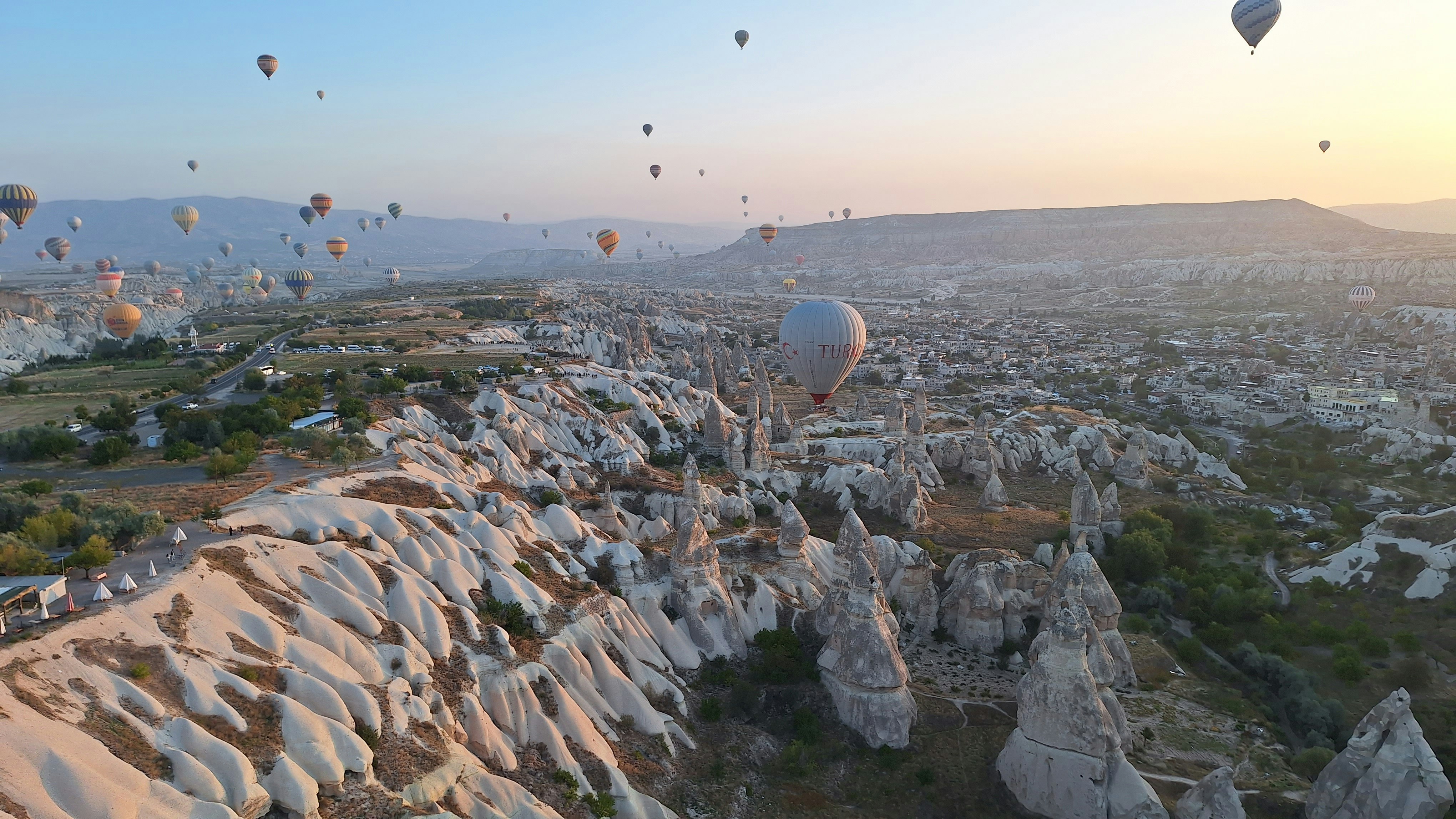 Hot air balloons drift over a rocky valley at sunrise, revealing fairy-tale formations and a village below. Wide shot emphasizes vast landscape with a calm, pastel sky.