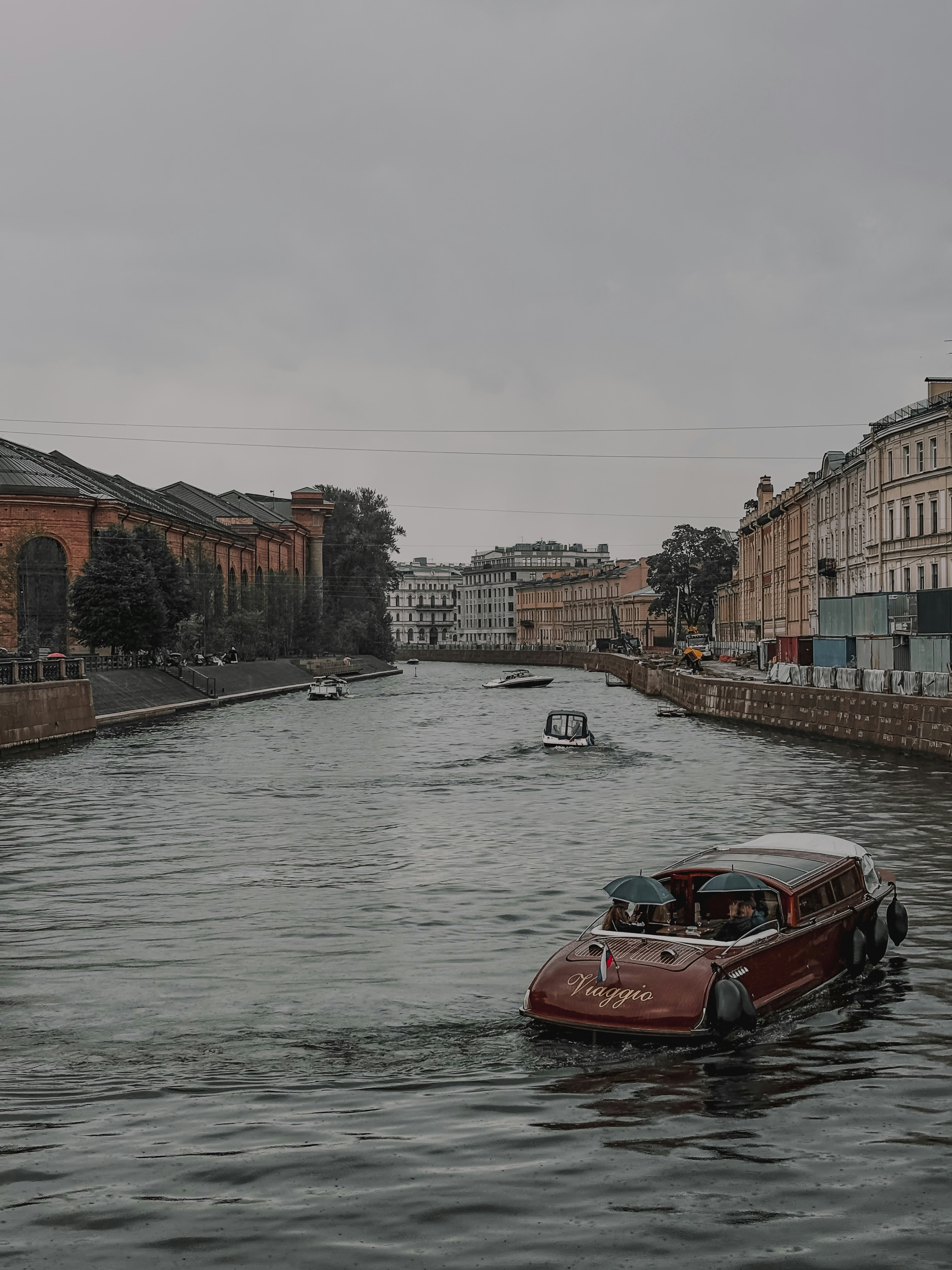 Red boat glides along a calm canal flanked by historic buildings and a stone embankment, capturing a tranquil urban waterway at dusk.