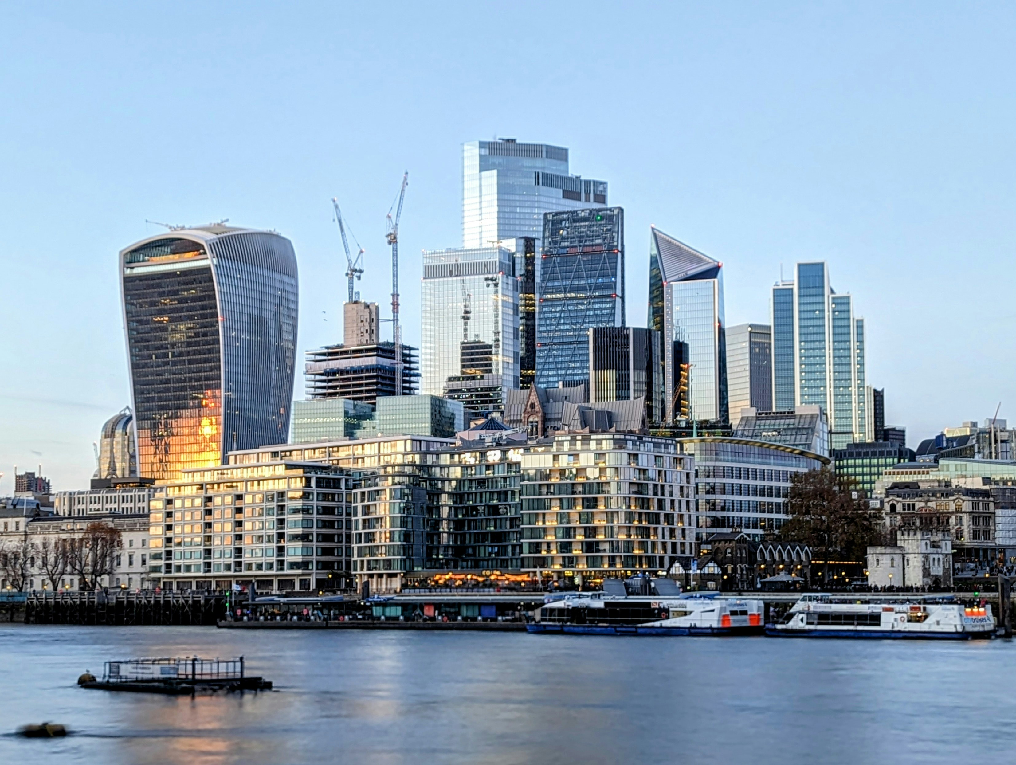 City skyline with modern glass towers reflecting sunlight along a riverfront, boats in the foreground and a mix of mid-rise buildings lining the water.
