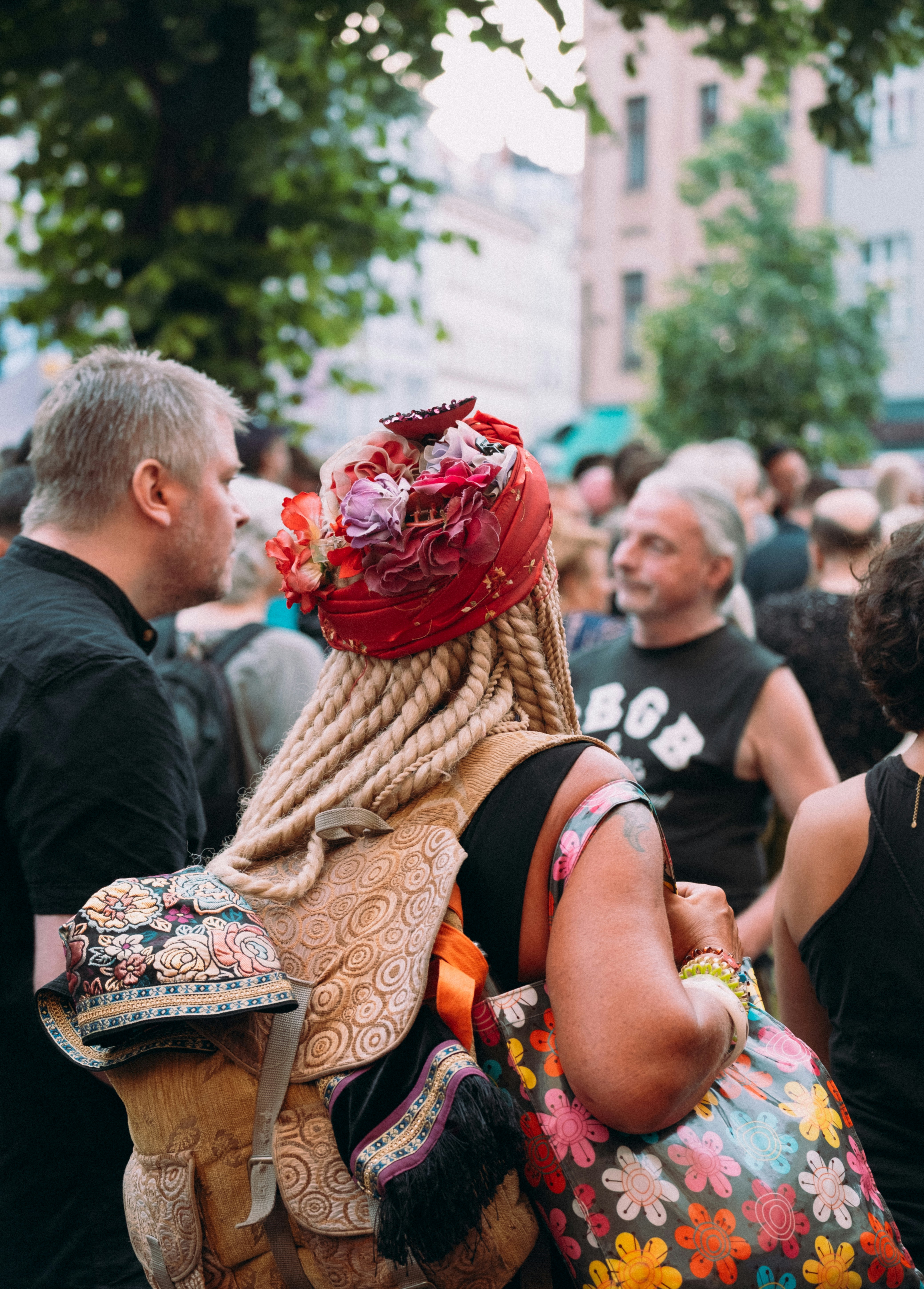 A vibrant woman with a red floral headscarf and braided hair stands among a crowd, carrying a floral tote bag and a patterned backpack in an outdoor gathering.