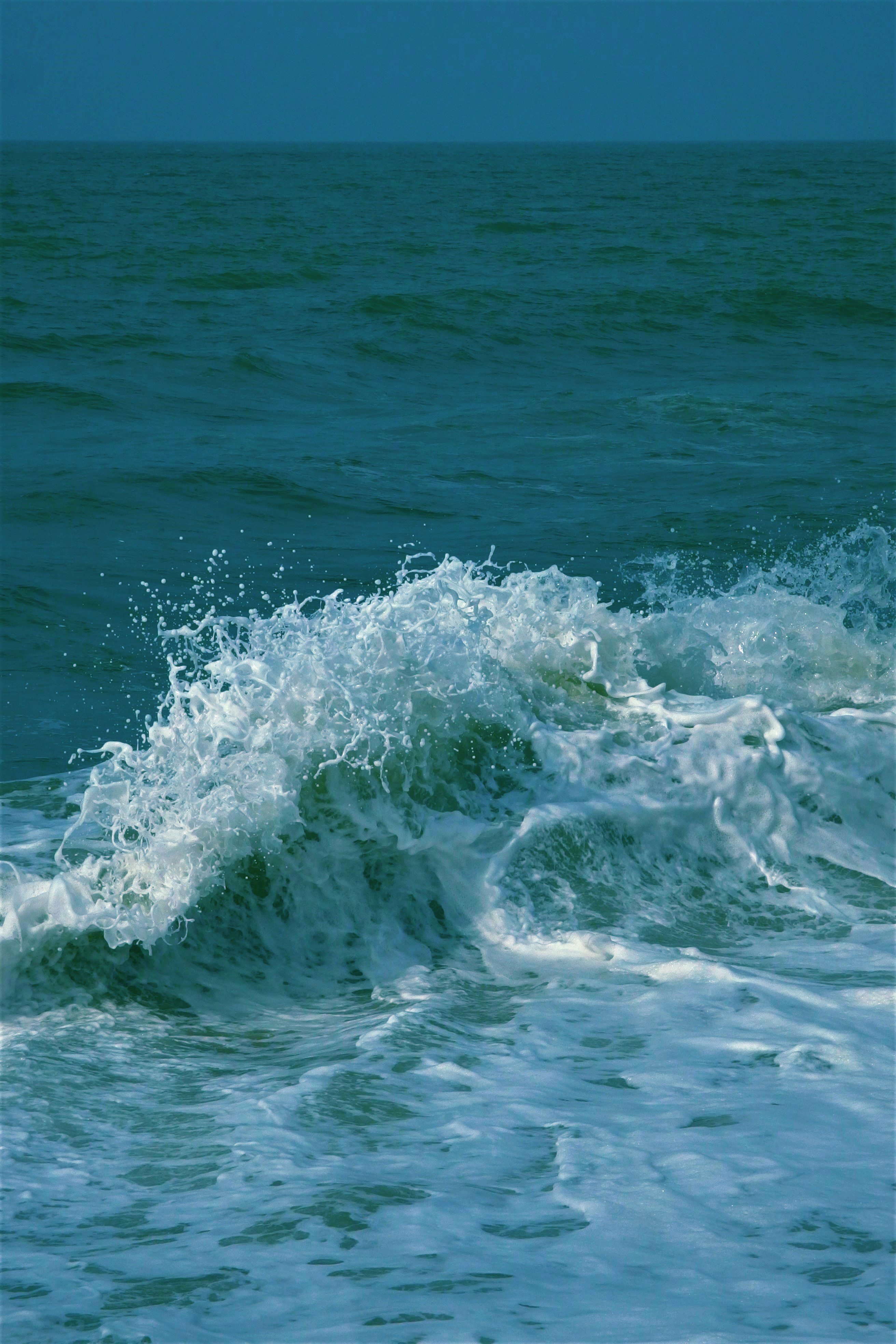 Vibrant sea wave breaking toward the shore with foamy spray and deep blue-green water.