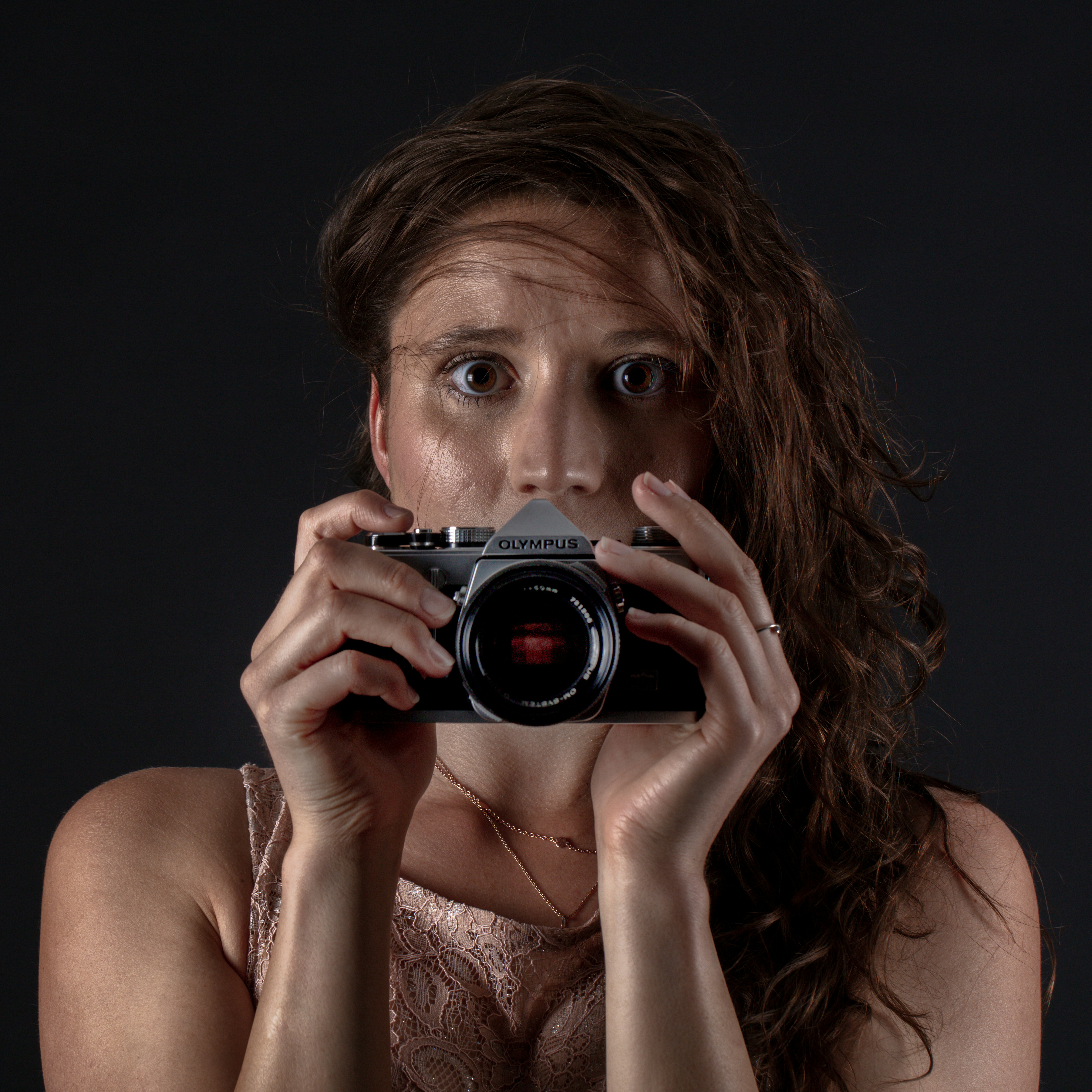 A woman holding a vintage camera up to her face against a dark background; the camera lens and upper torso are visible, hinting at a portrait session or photographic moment.