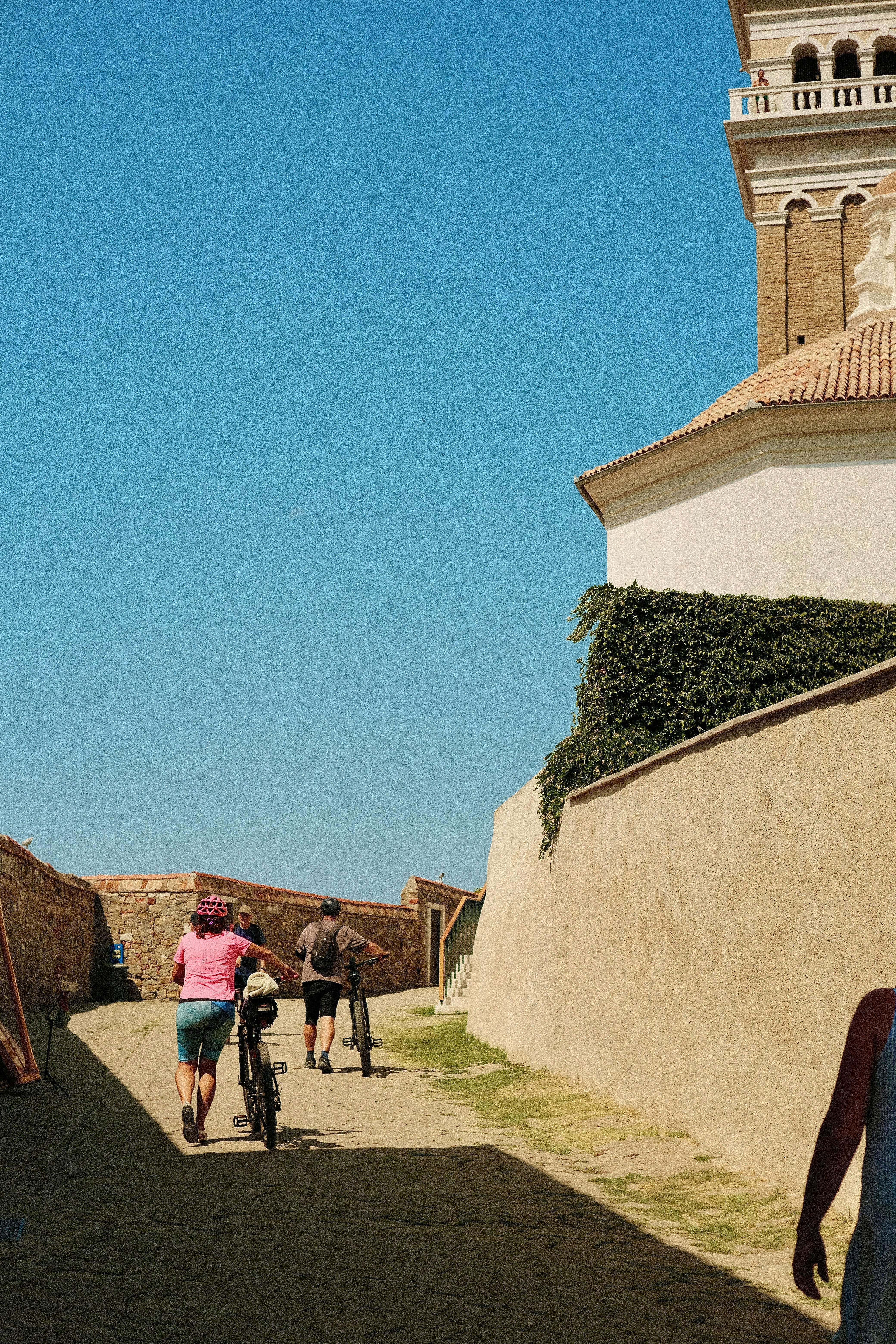 Sunlit path beside ancient walls carries cyclists toward a sunlit courtyard, with a tower peeking above and ivy trailing the wall.