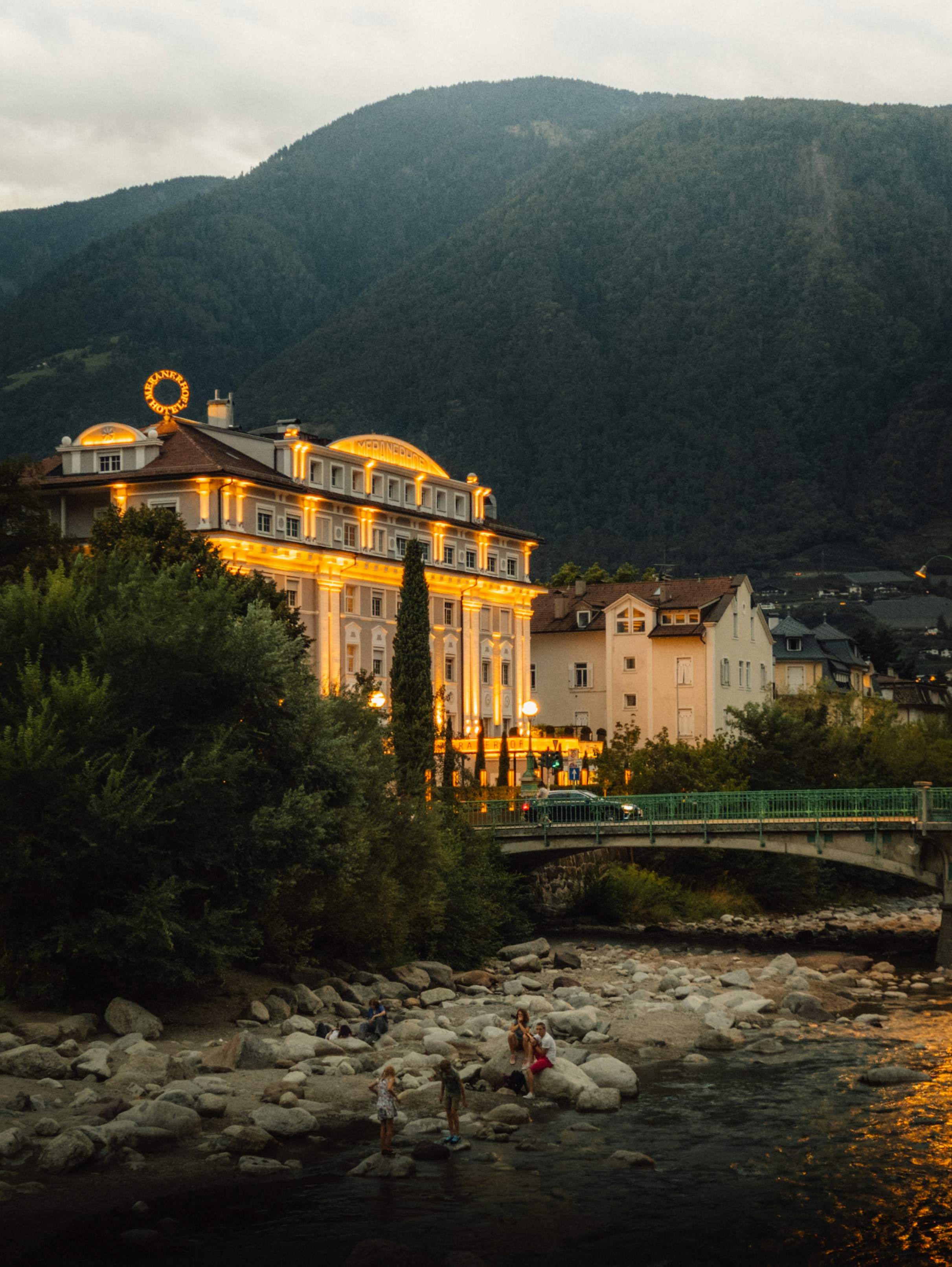Illuminated hotel sits by a rocky river with a footbridge and surrounding village at dusk; warm lights contrast with dark mountain backdrop. A small group of people gathers near the water’s edge.
