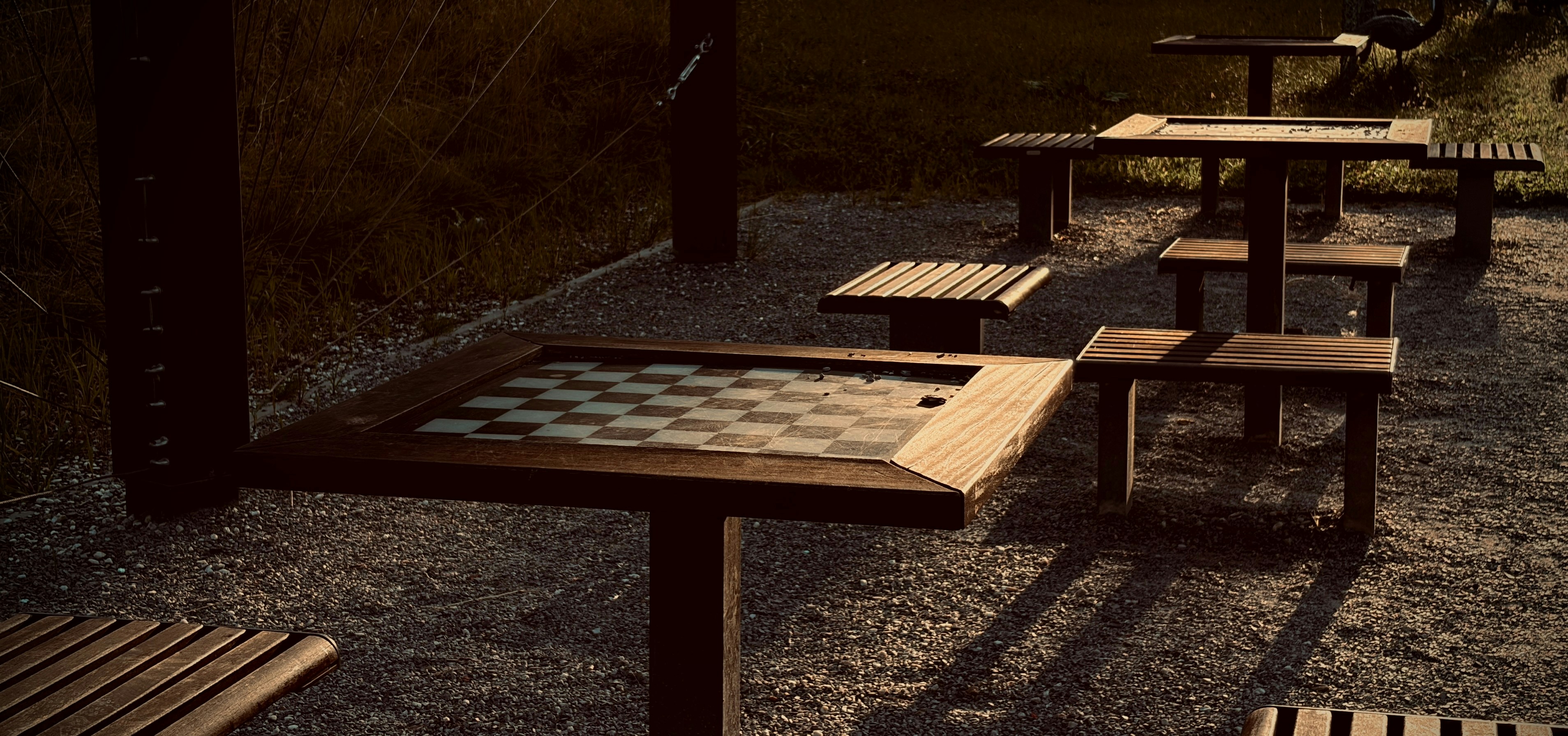 Muted park scene with wooden chessboard-style table and benches casting long shadows on gravel, empty and still at dusk.